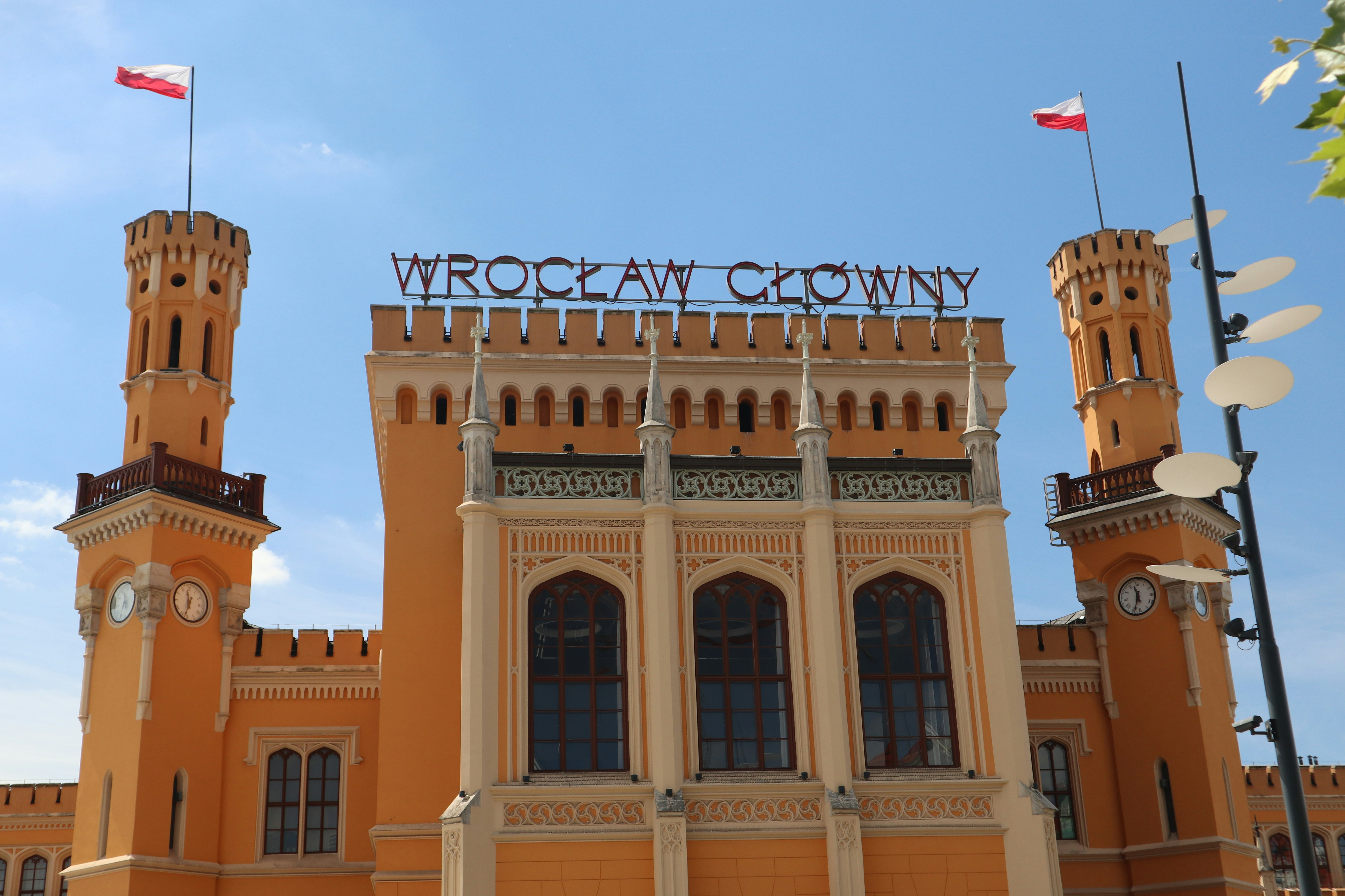 Bright orange train-station style building with twin cylindrical towers and decorative central facade, under a clear blue sky.
