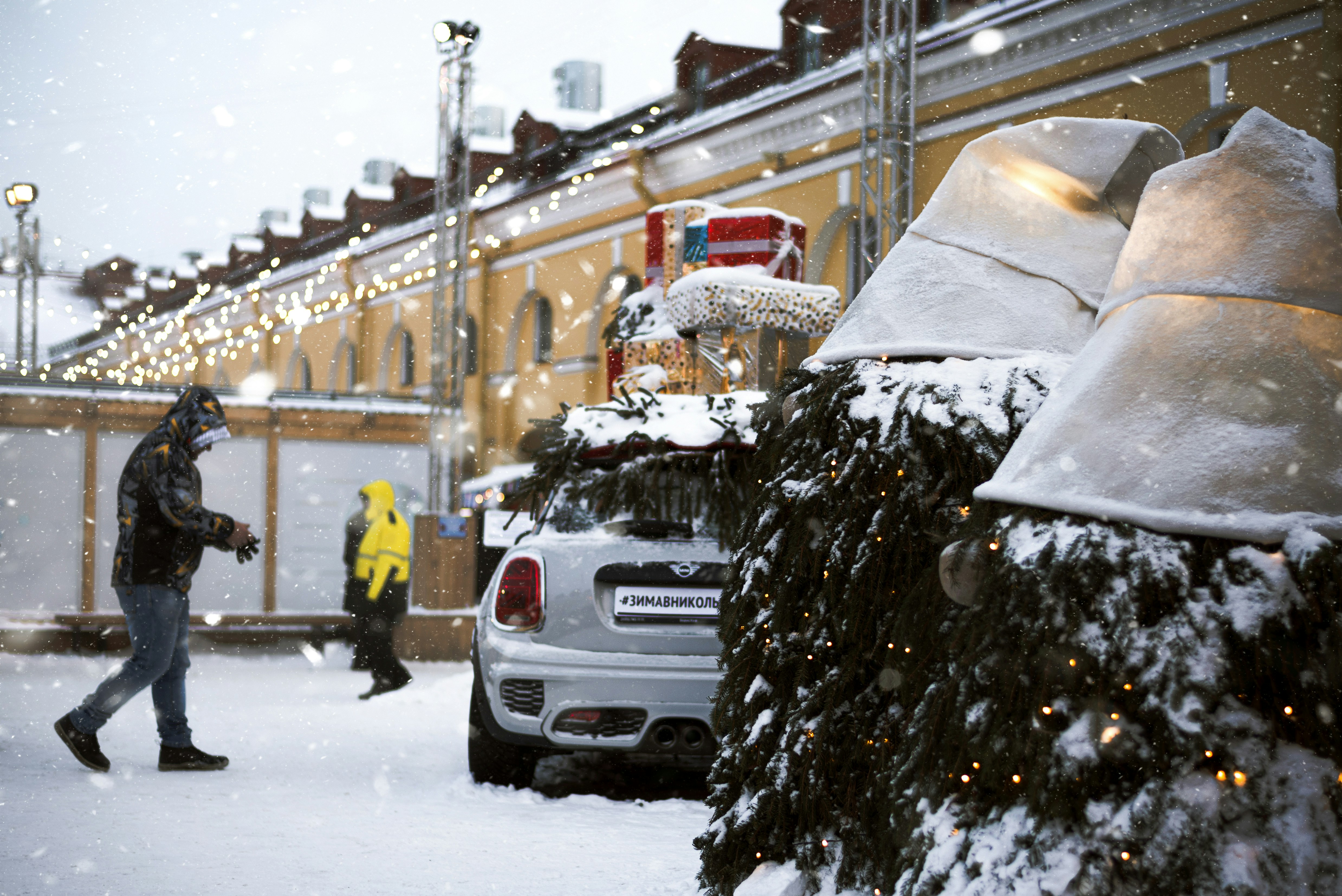Car parked on a snowy street with festive lights and pedestrians in winter clothing.