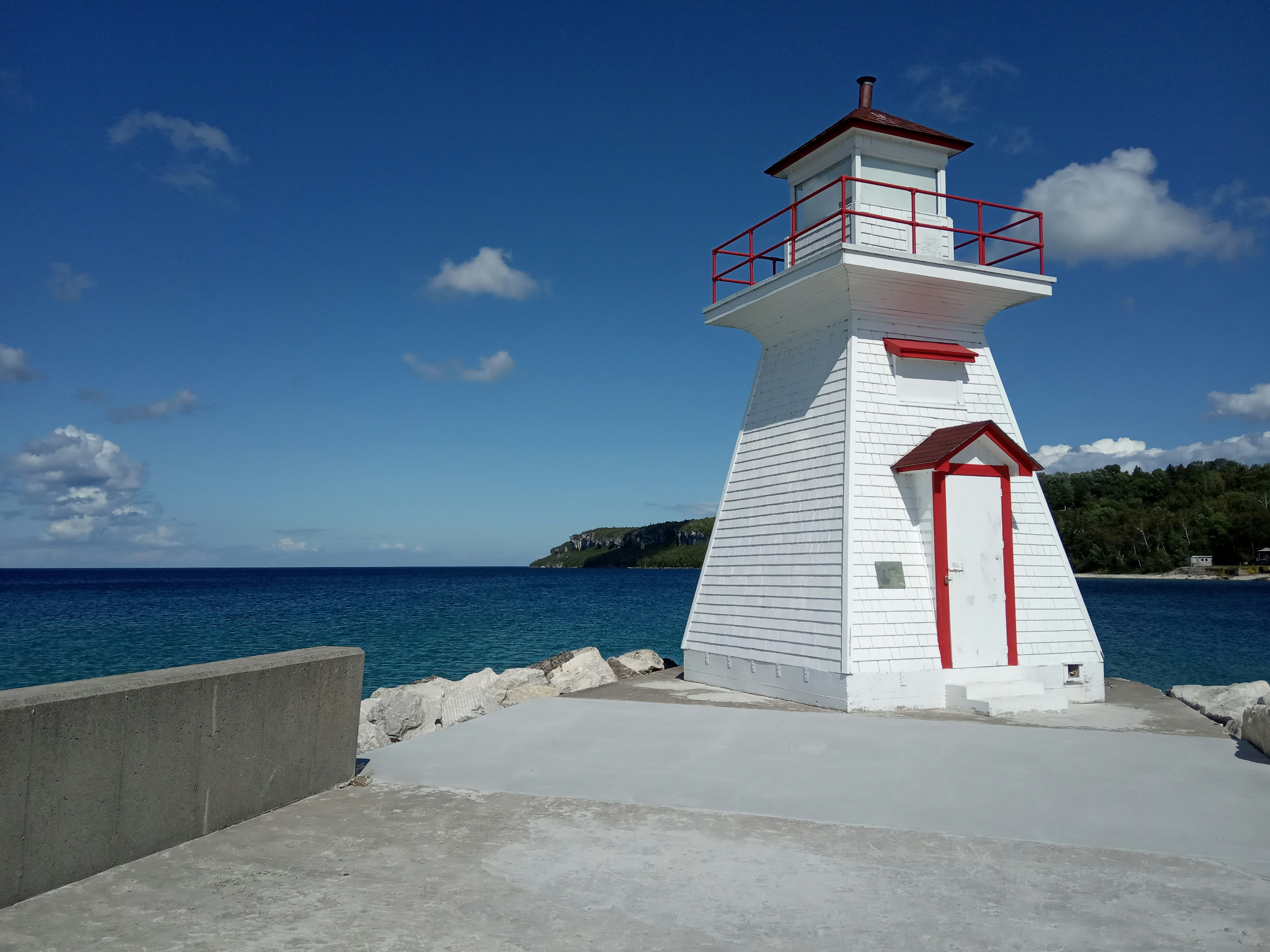 White lighthouse sits on a concrete pier beside calm blue water under a clear sky, captured in a crisp photograph.