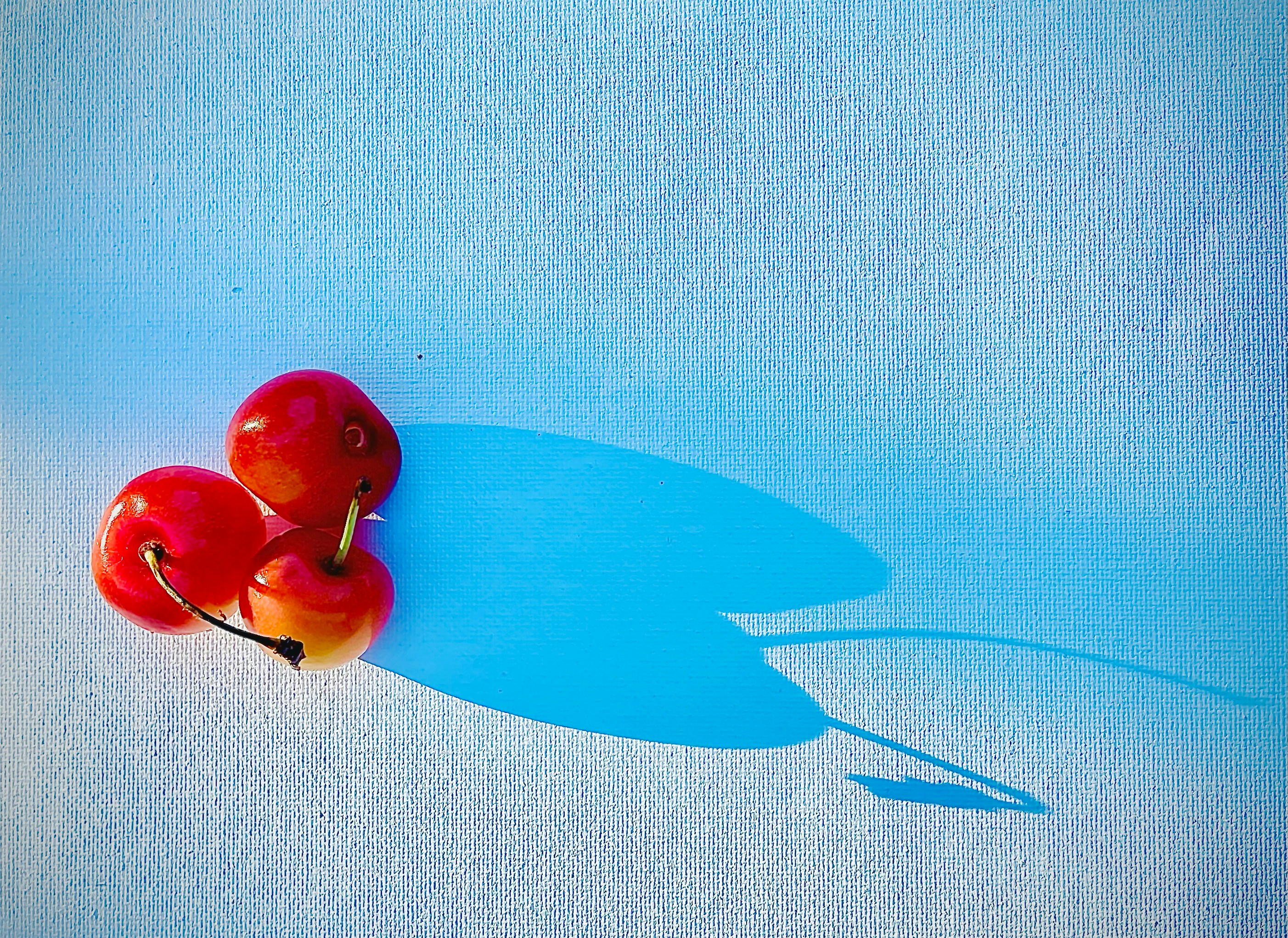Two bright cherries cast a bold blue shadow across a pale canvas