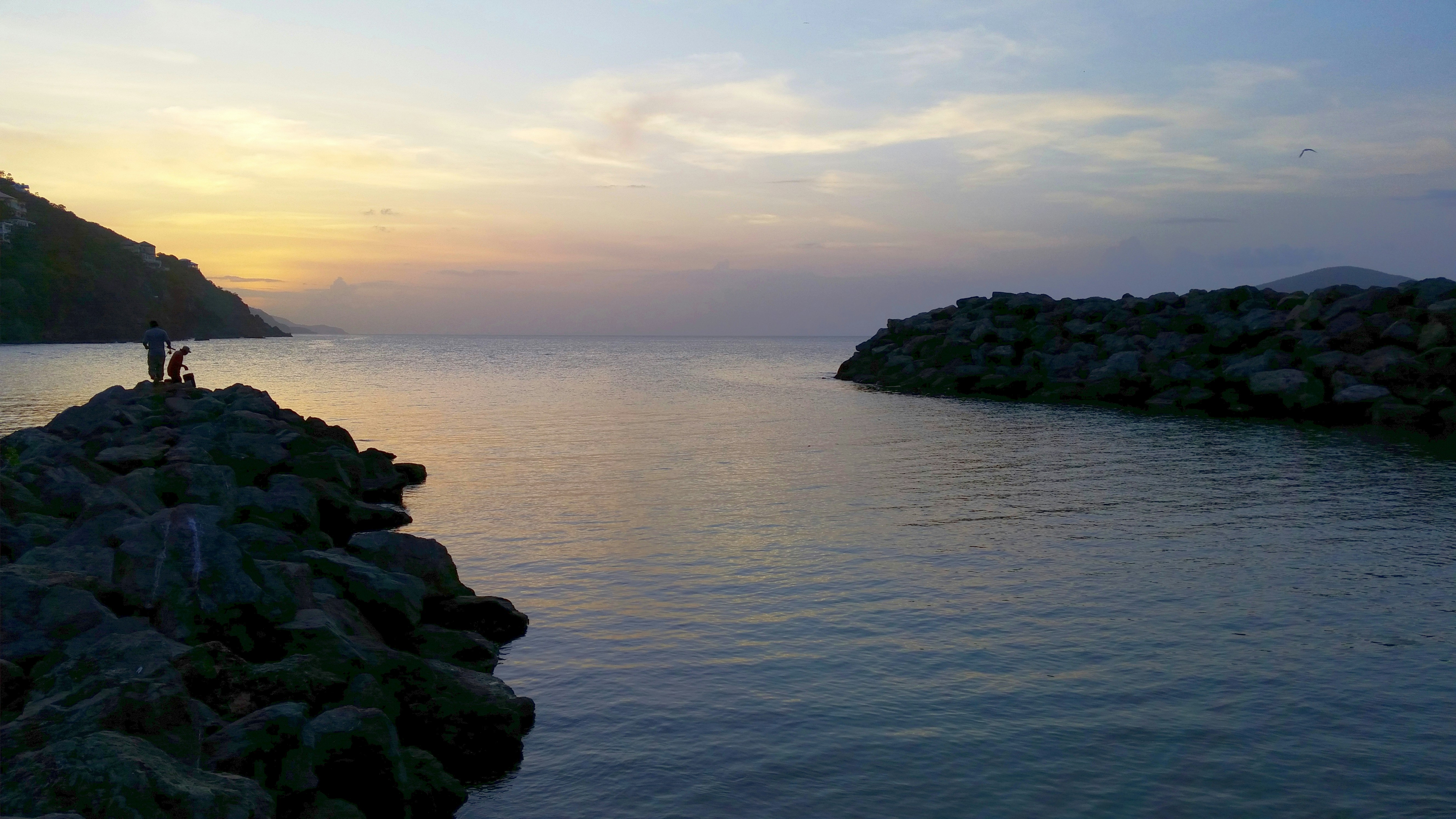 Two silhouettes stand on a rocky jetty as the sun sinks, casting warm pastel light over calm water. A seascape photograph capturing dusk by the shore.