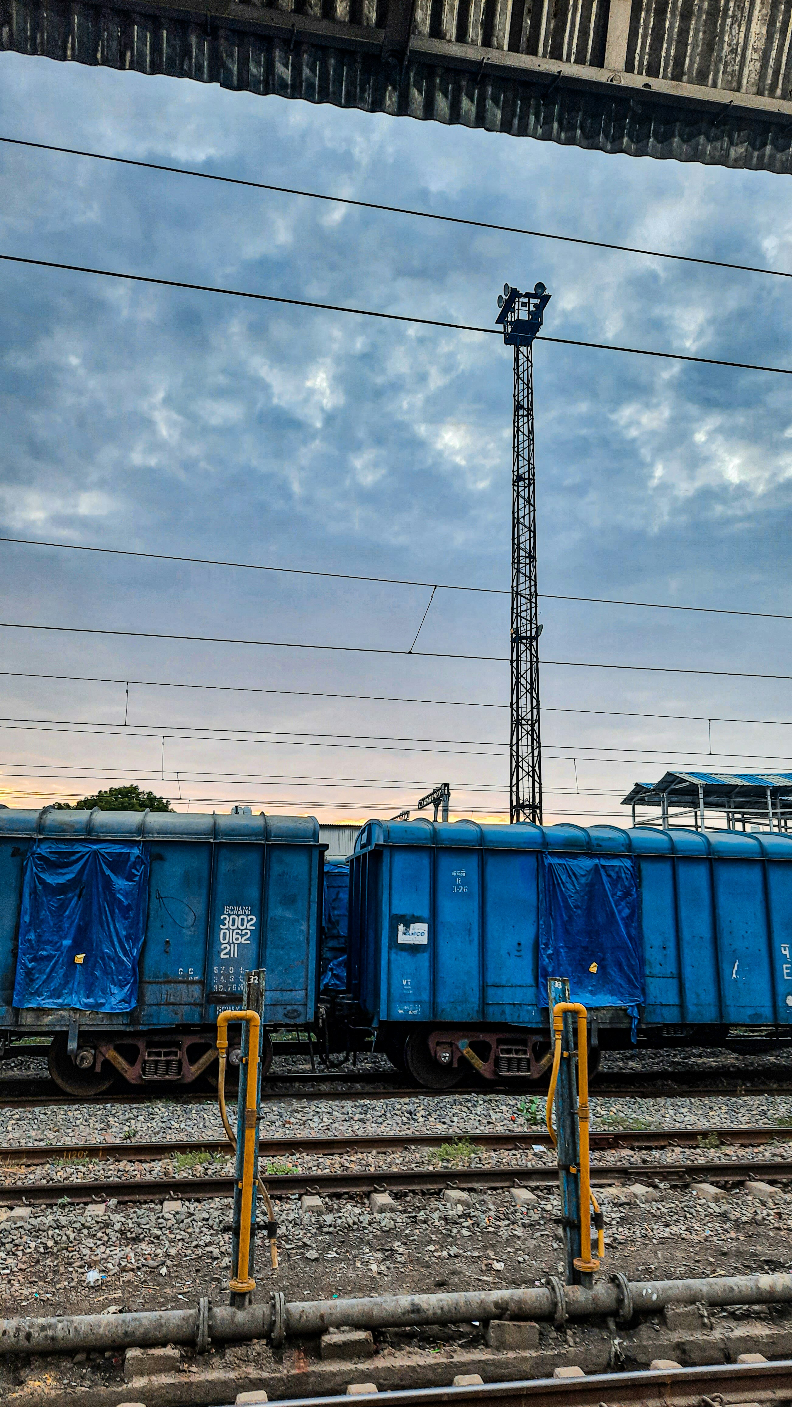 Blue freight wagons sit along tracks beneath overhead power lines, with a tall lattice tower rising against a cloudy sky.
