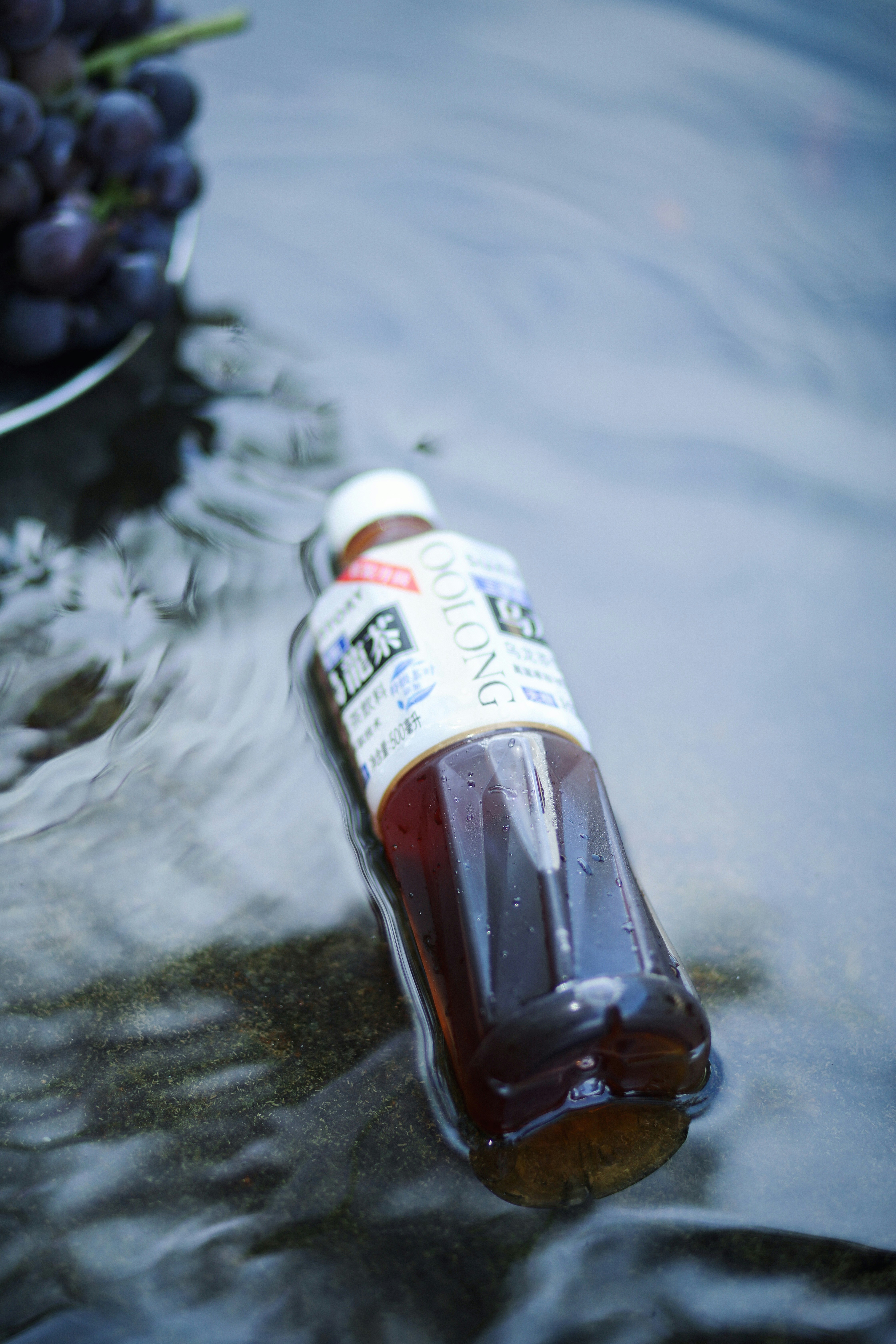 A bottle labeled Oolong rests partially submerged in a shallow, reflective pool, with blurred grapes nearby.
