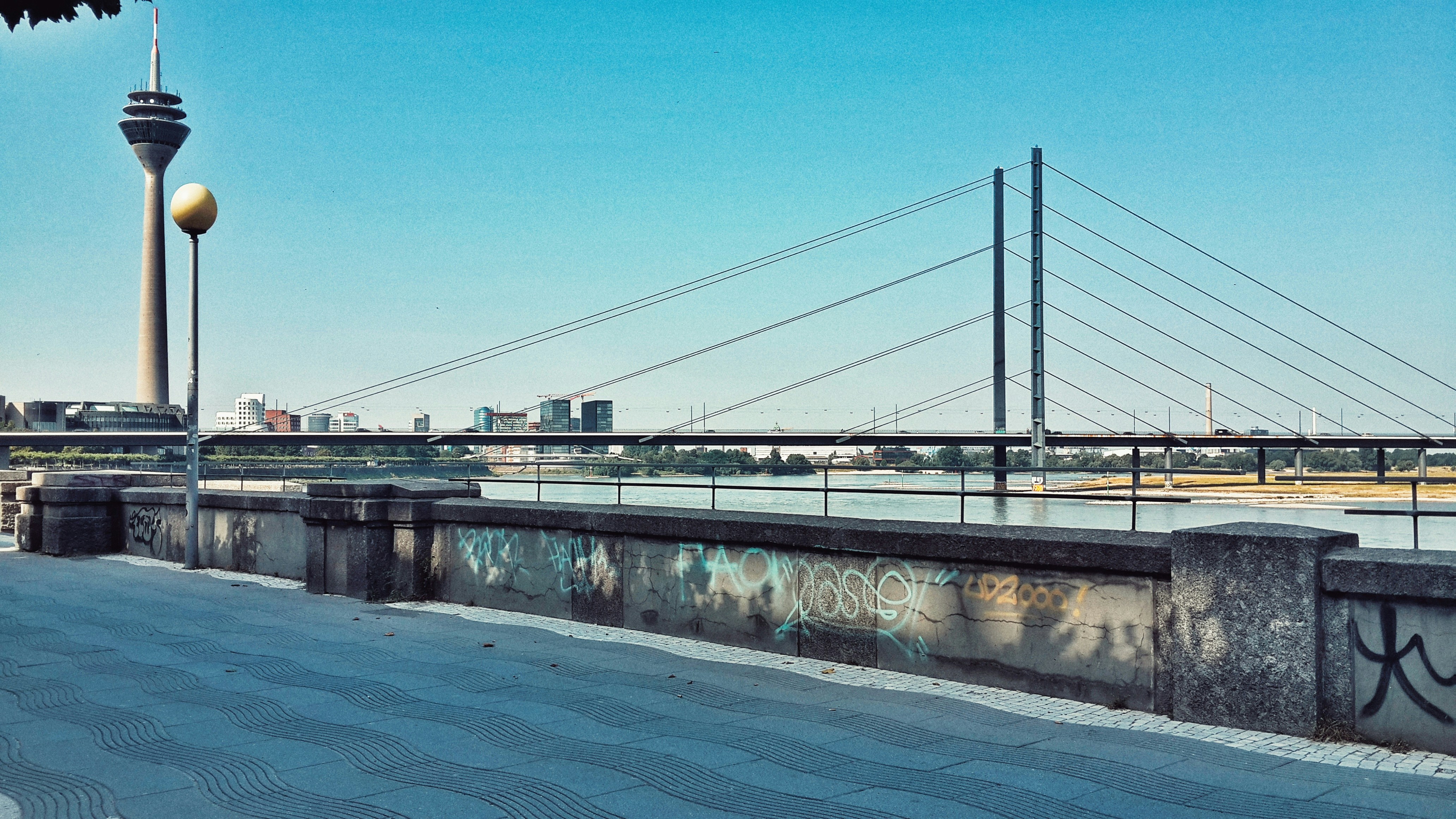 City riverfront promenade with graffiti on a concrete barrier, a distant suspension bridge, and a distinctive tower with a spherical top on the left. This daytime urban photograph captures the calm riverside scene and architectural silhouettes.