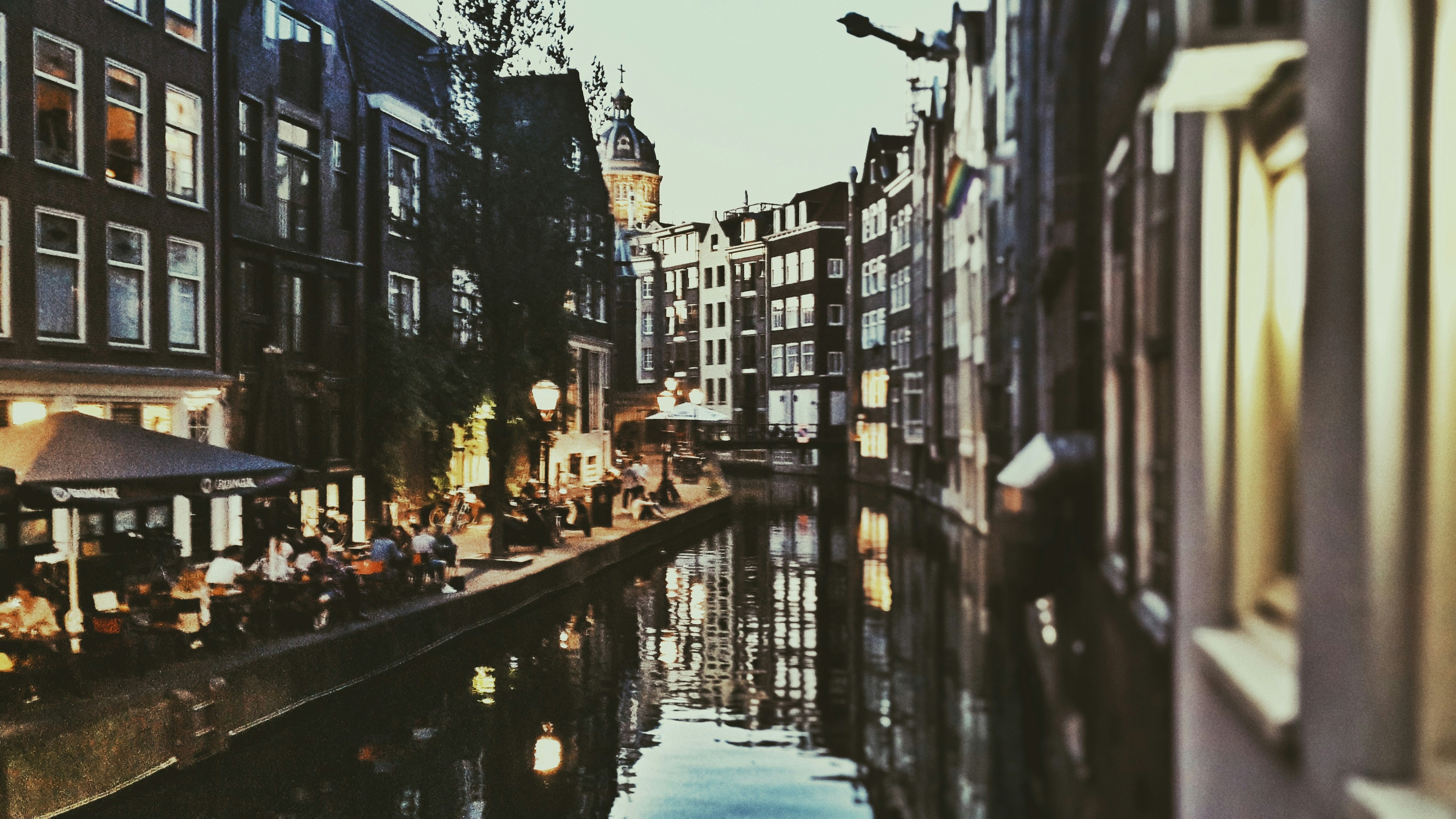 Nighttime canal street with warm lamp glow reflecting on the water, flanked by narrow gabled buildings. A distant tower anchors the horizon along the quiet canal.