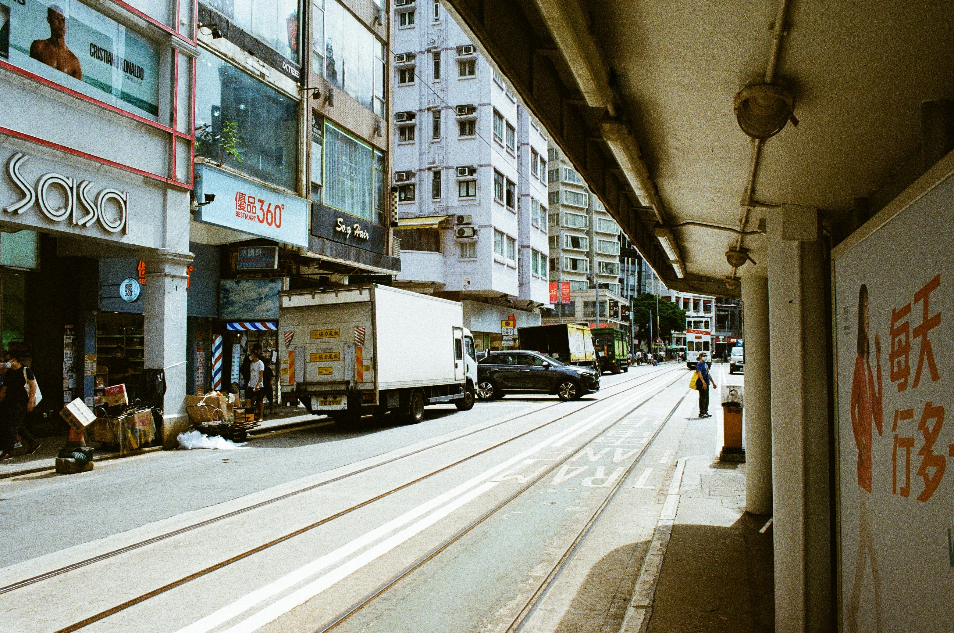 City street with tram tracks, lined by buildings and parked trucks under a cloudy sky.
