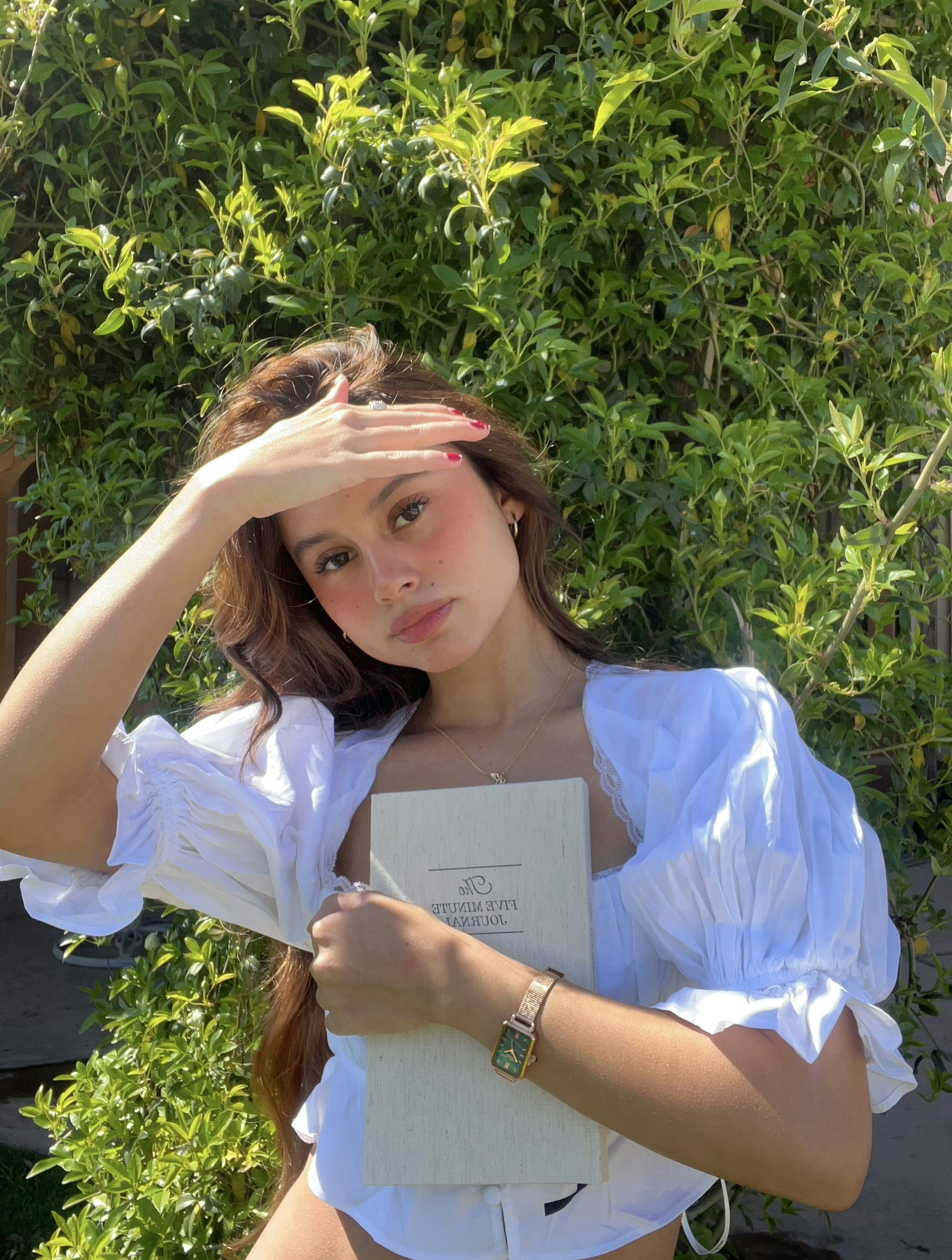 A young woman in a white blouse shields her eyes from bright sun. She holds a light notebook close to her chest as green foliage forms a lush backdrop.