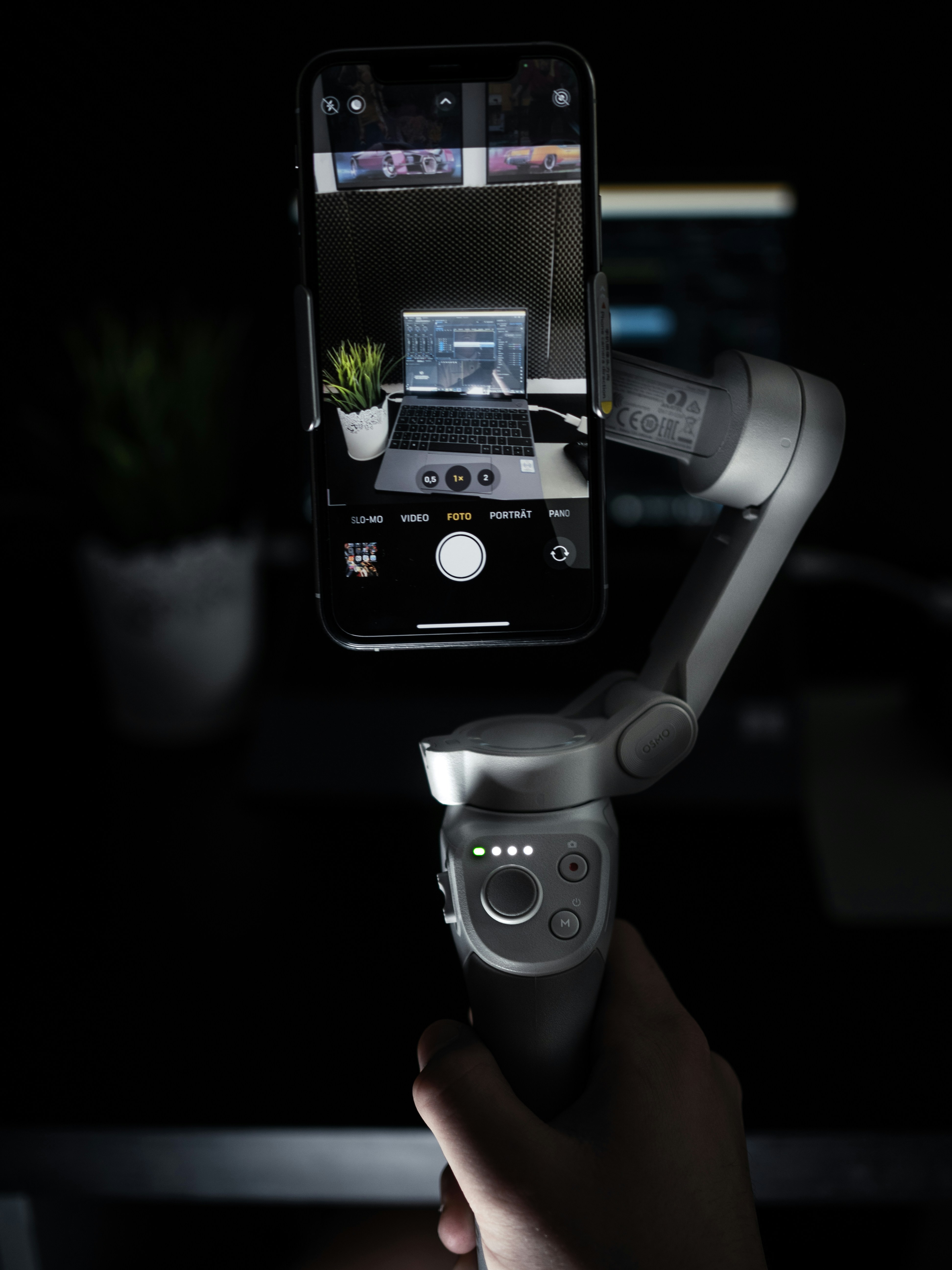 A smartphone mounted on a gimbal captures a digital workspace featuring a laptop and a potted plant, illuminated against a dark backdrop.