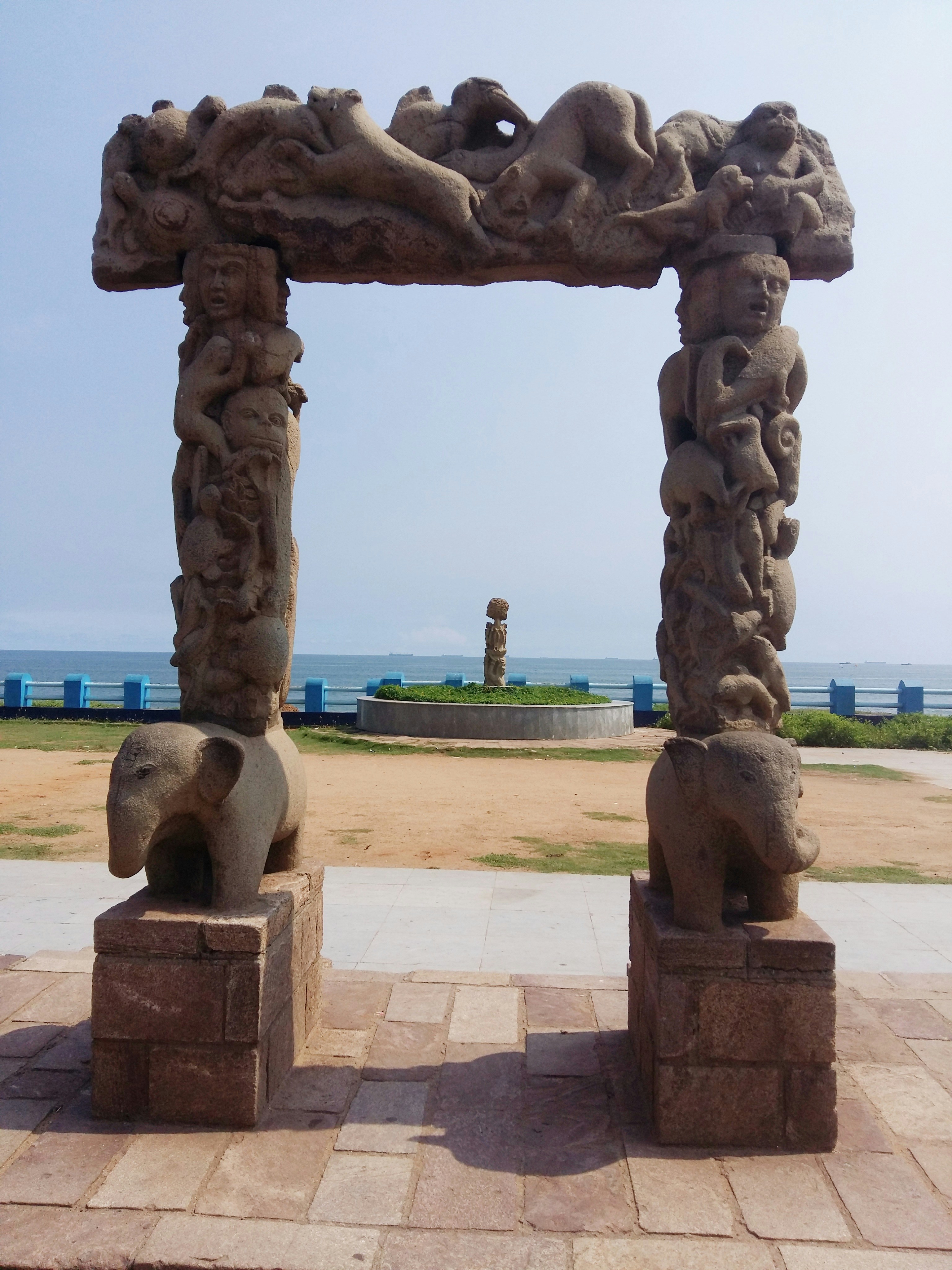 Intricately carved stone archway featuring figures and elephants, leading to a serene coastal view. The scene captures a blend of artistry and nature.