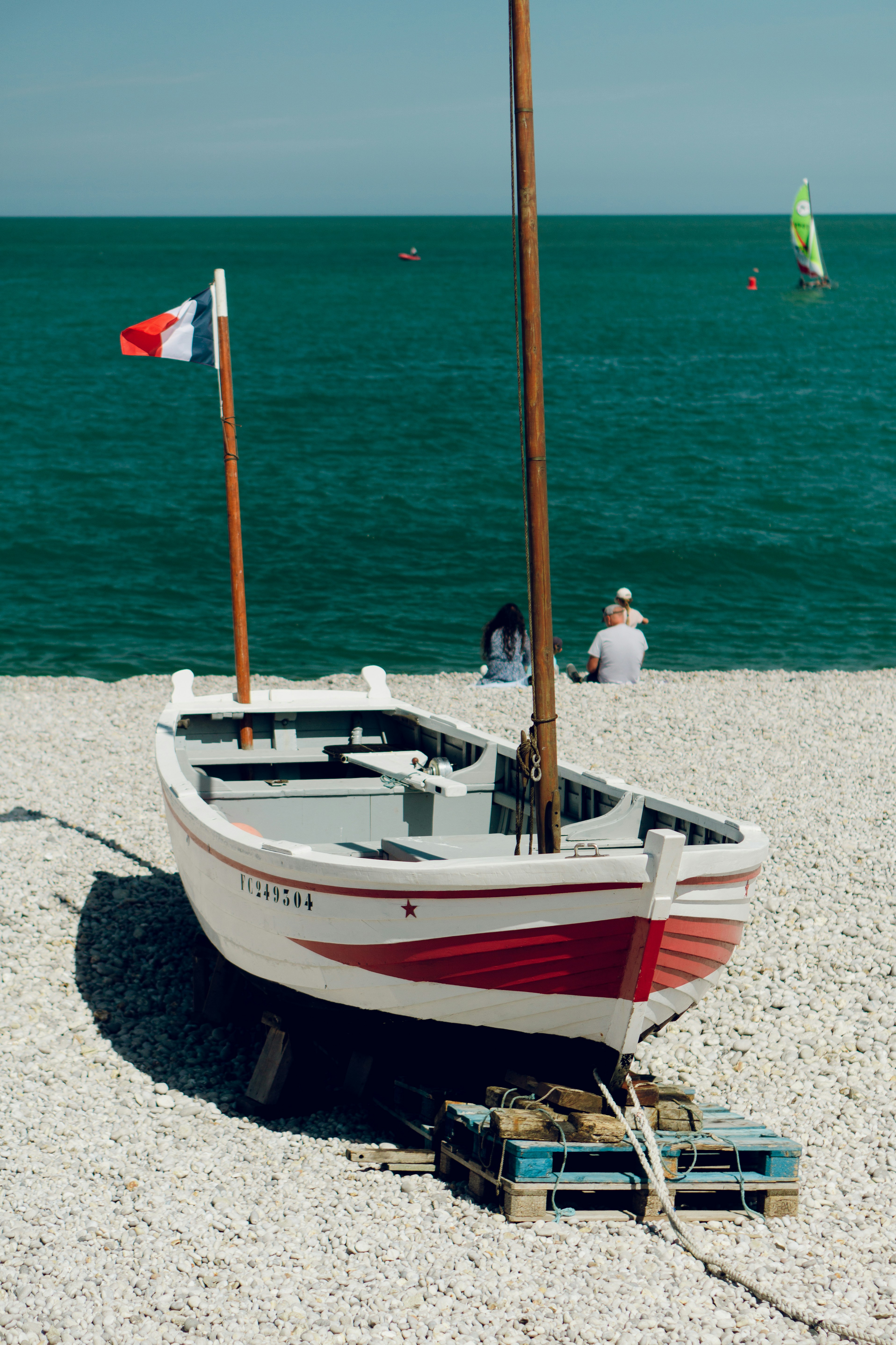 Traditional fishing boat resting on pebbled beach with a French flag, while people enjoy the seaside in the background.
