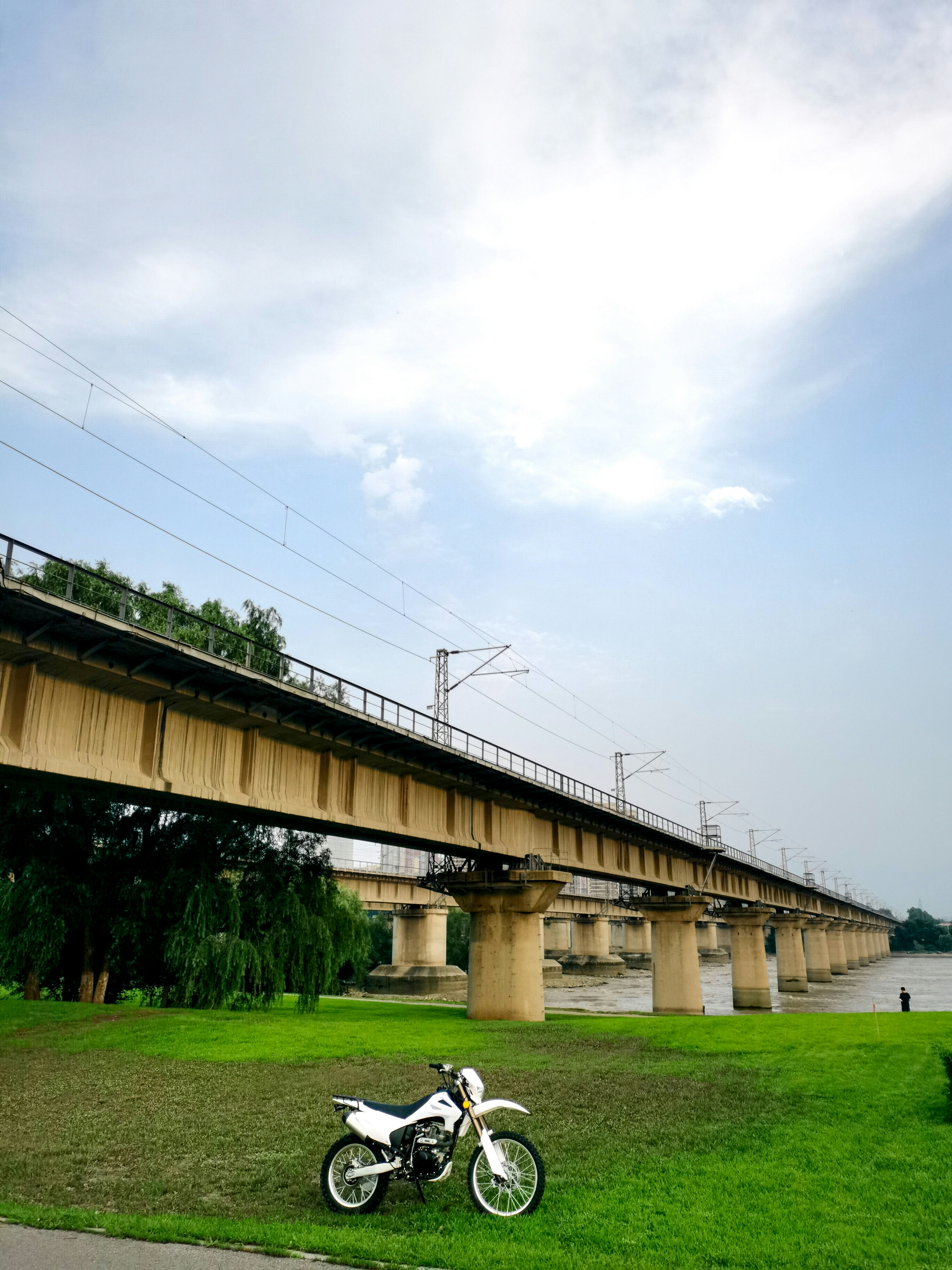 A white dirt bike rests on a grassy patch beneath a long concrete railway viaduct, with a bright sky and a distant figure along the tracks.