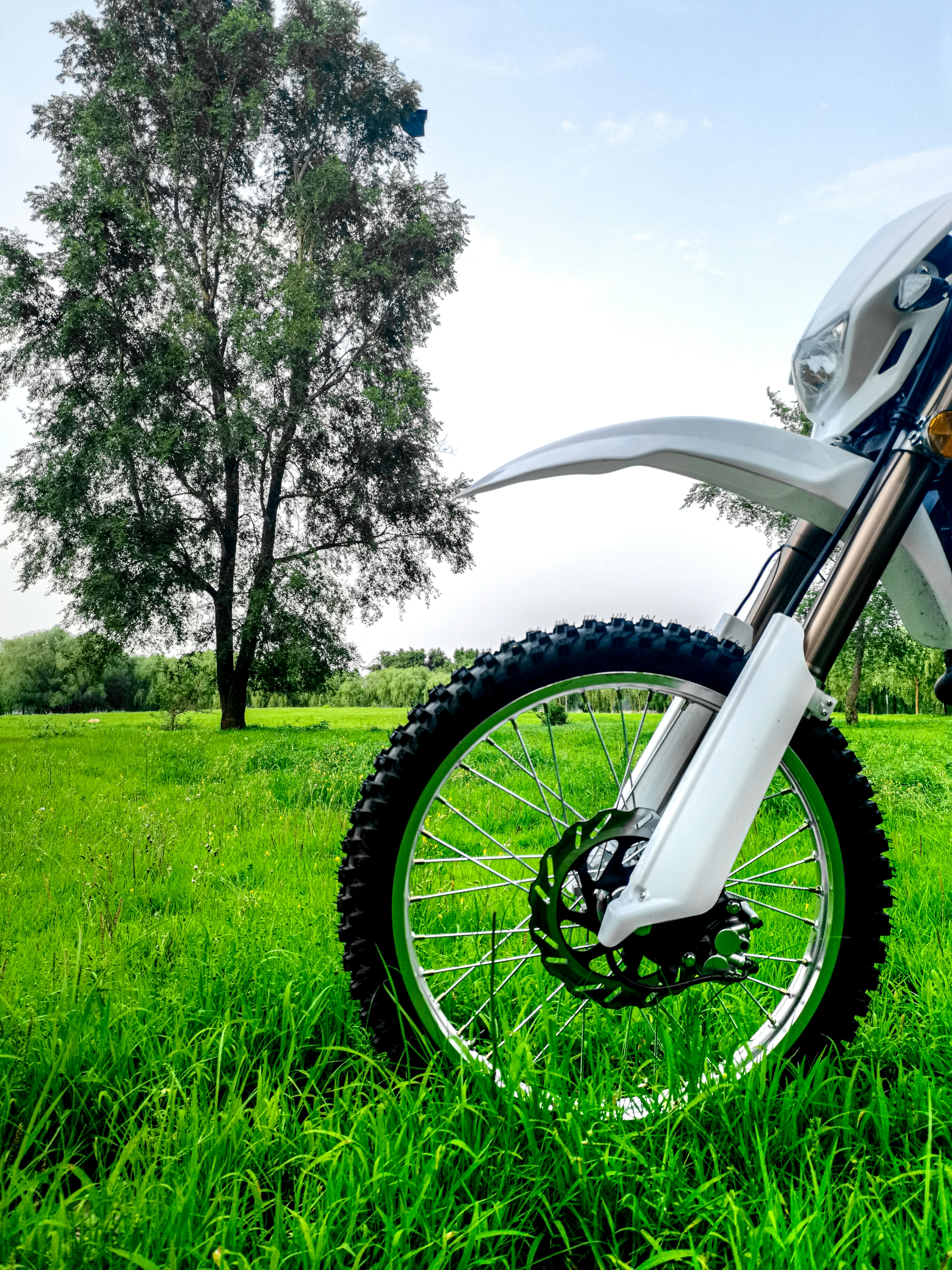 Close-up of a dirt-bike front wheel and fork resting on bright green grass, with a lone tree and blue sky in the background.