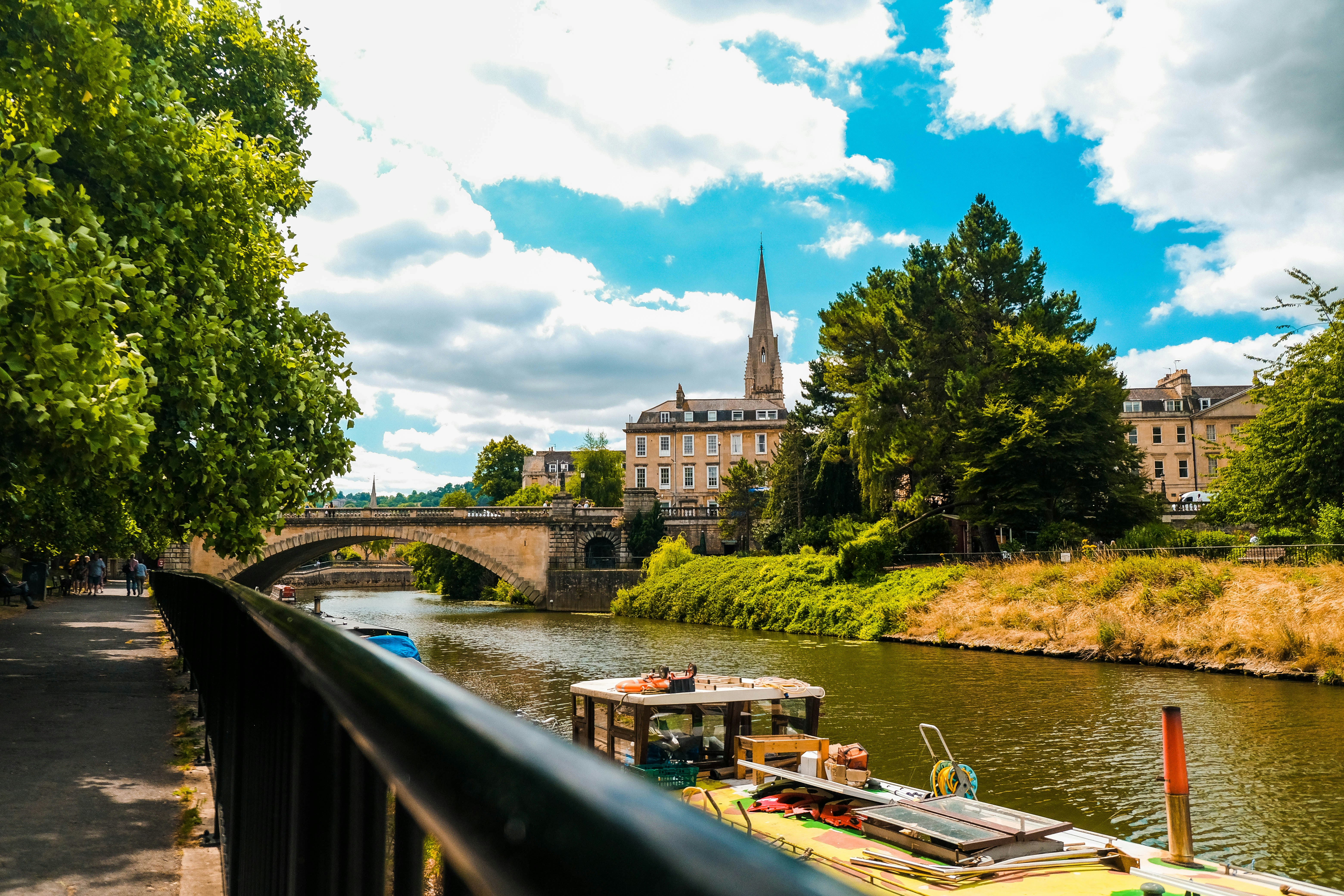 Boats float on a river beneath a stone bridge, with historic buildings and a church spire in the background under a vibrant sky.