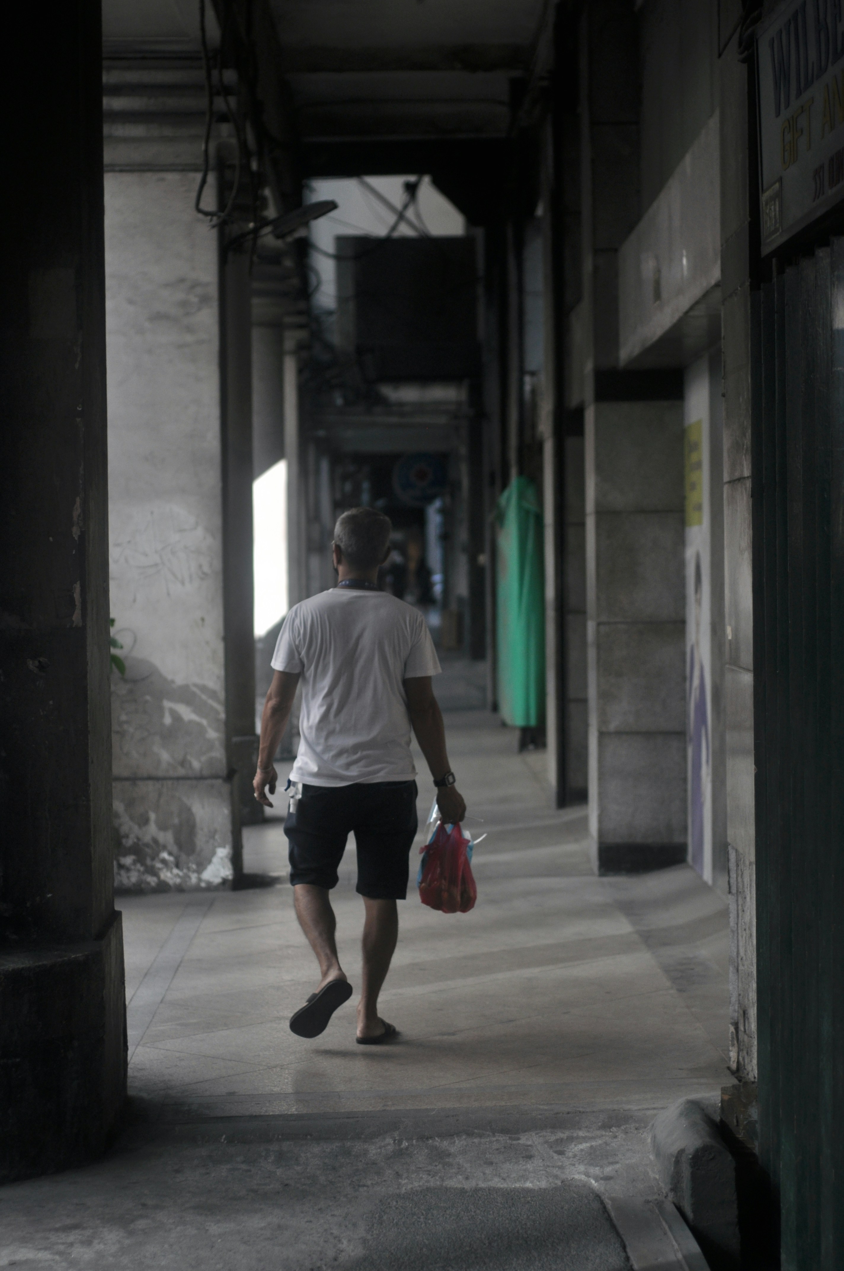 A lone figure walks through a dimly lit corridor, carrying a bag, with shadows casting an atmospheric depth. The scene captures urban solitude and quiet contemplation.