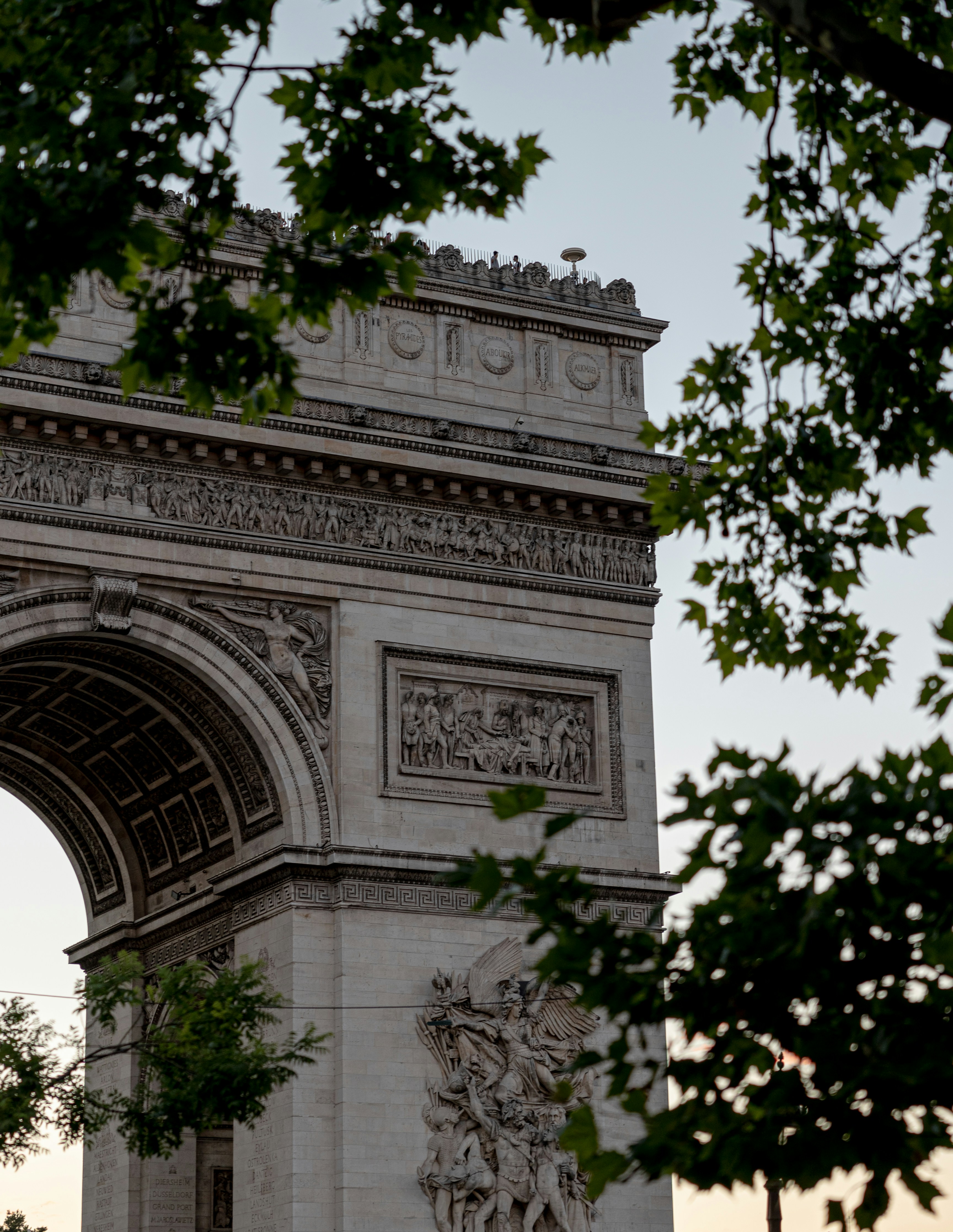 Whispering Carvings: Arc de Triomphe Framed by Dappled Leaves at Dusk