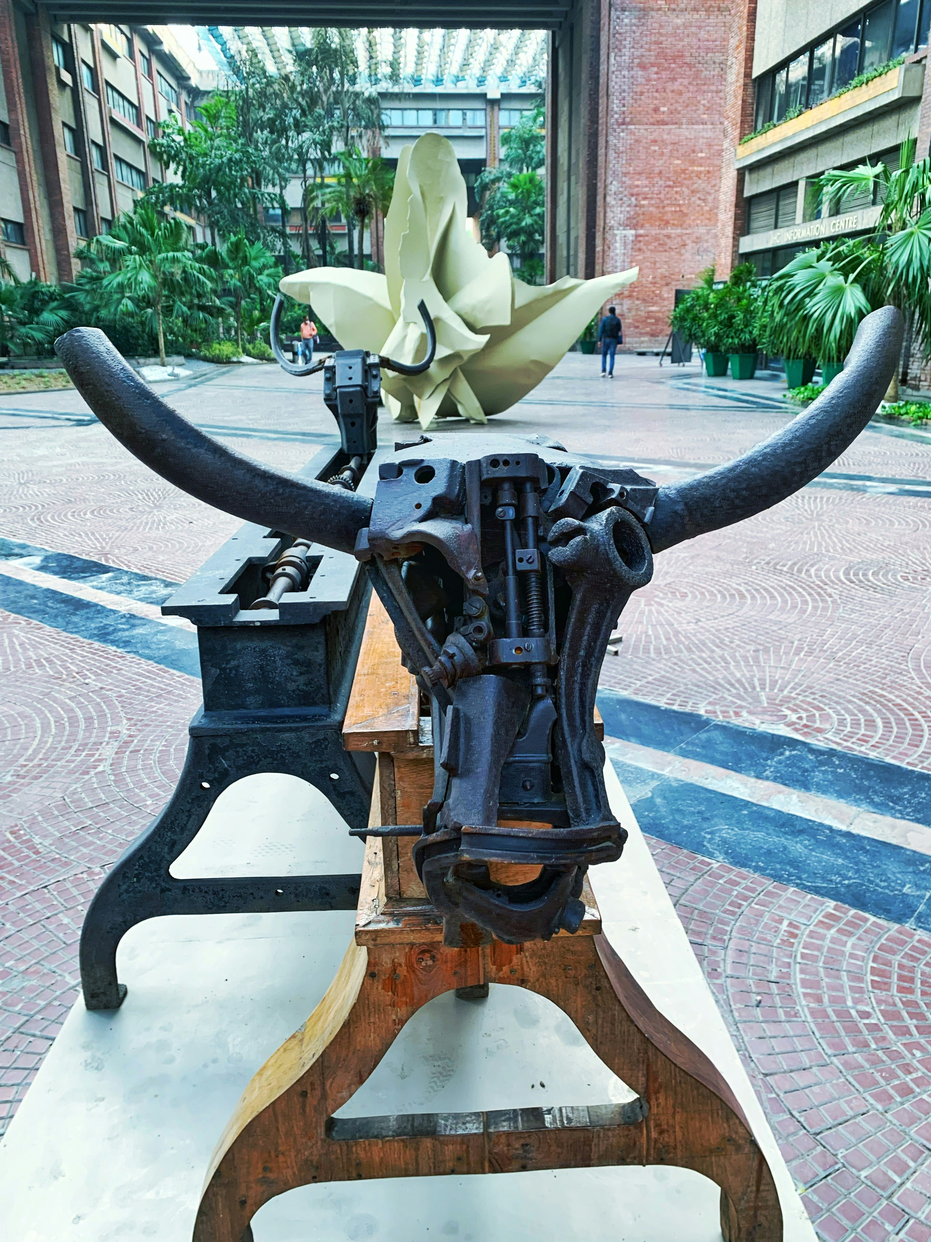 Sculpture of a bull's head crafted from metal components, positioned on a wooden base amidst a lush courtyard with greenery and modern architecture.