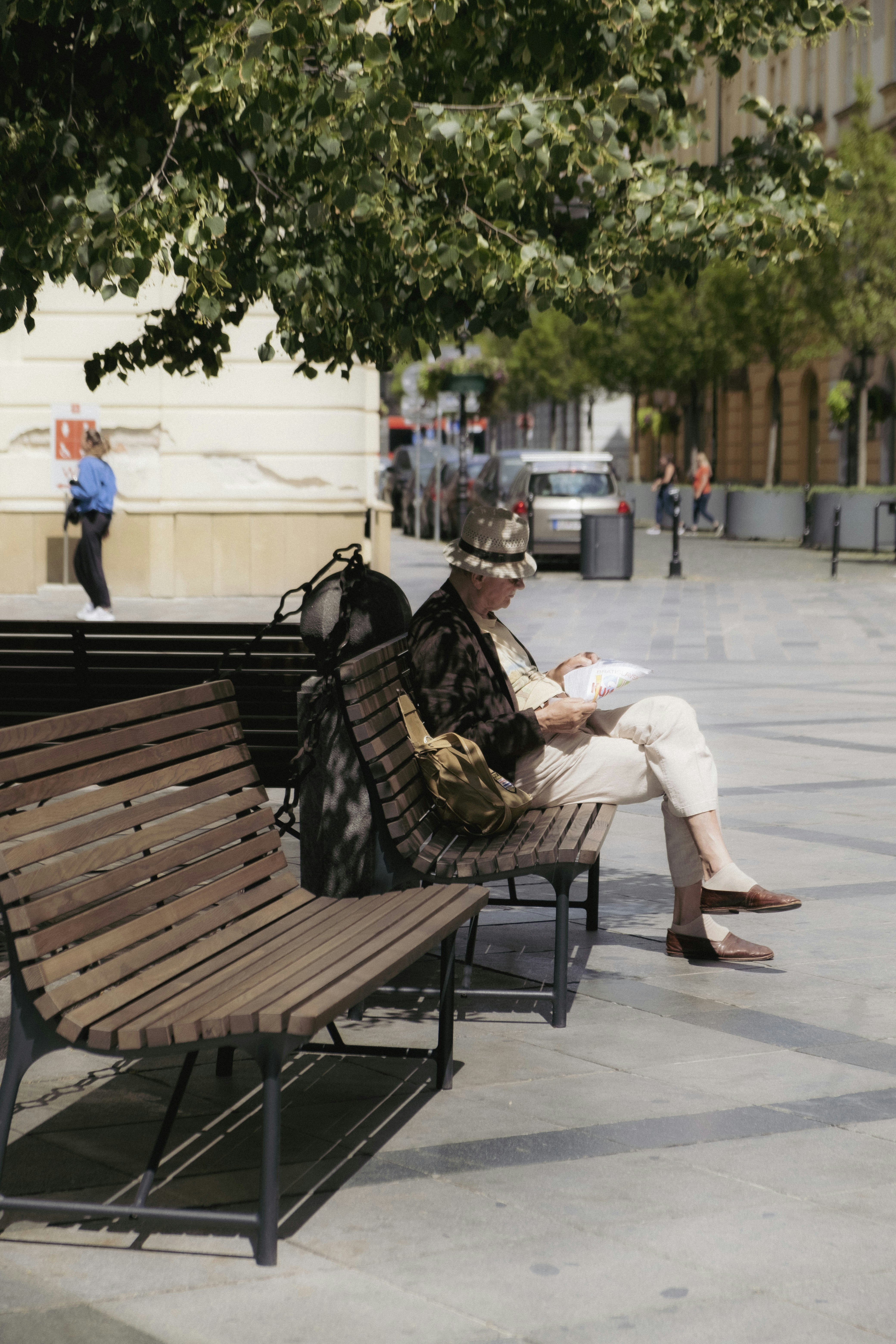 Elderly person seated on a park bench, engrossed in reading, surrounded by a lively city backdrop. The scene captures the essence of urban tranquility.