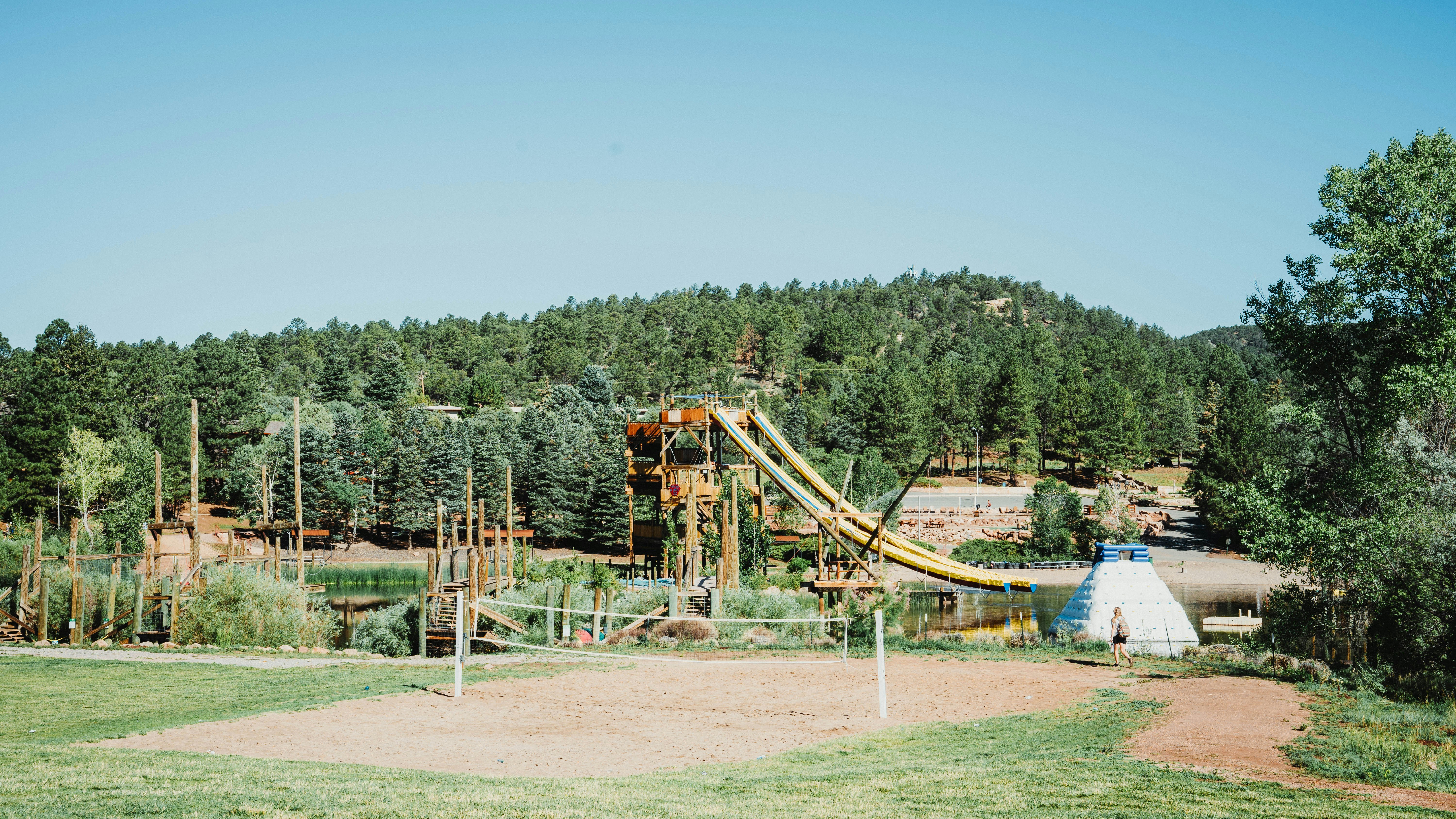 A vibrant water slide cascades down a hillside surrounded by lush greenery, with a sandy volleyball court in the foreground. The scene captures the essence of outdoor fun and adventure.