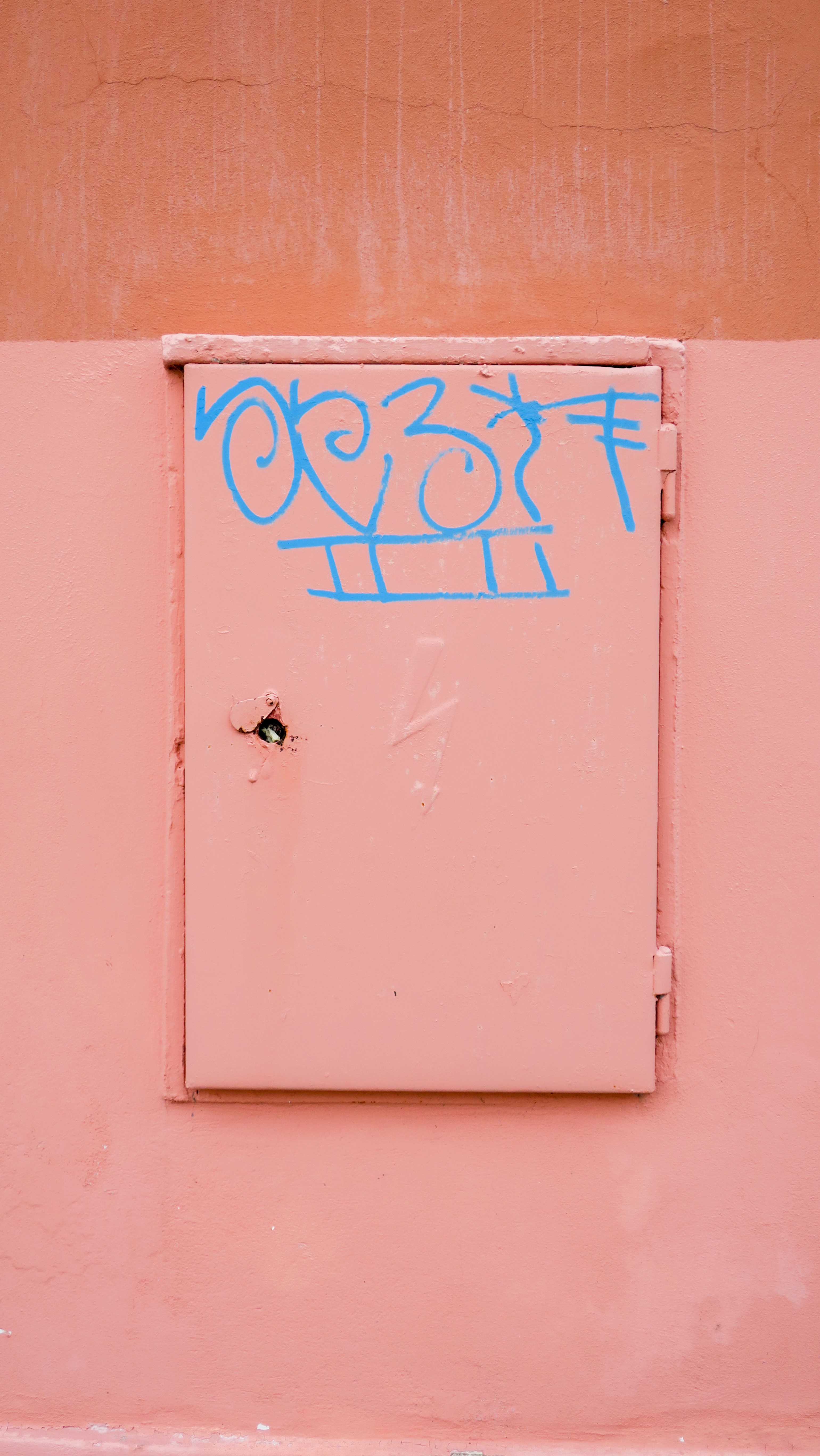 A pink wall featuring a blue graffiti-covered access panel, displaying a blend of urban art and muted tones.