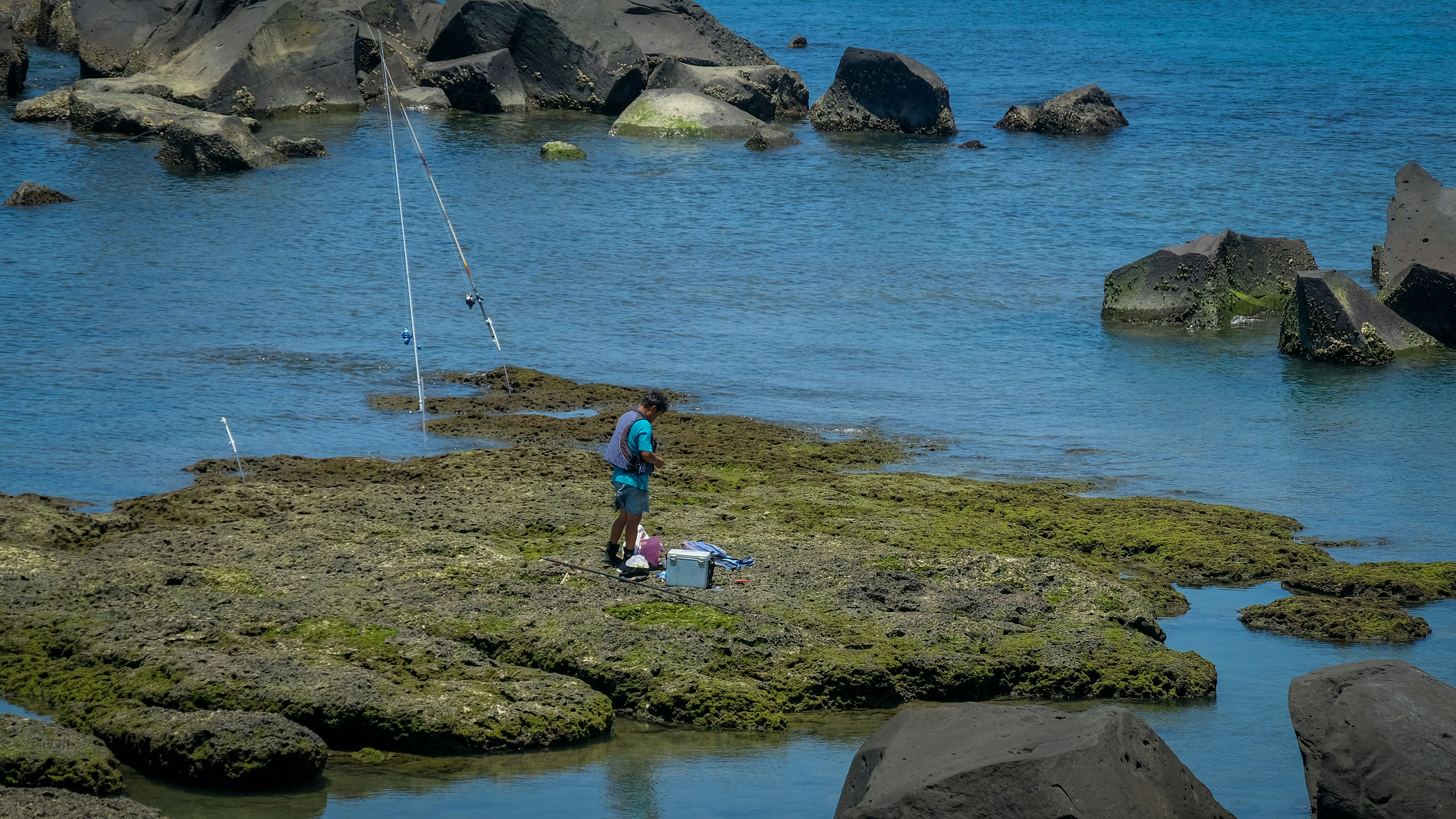 Fisherman stands on rocky tide pools, preparing gear amidst calm blue waters and scattered boulders. A fishing pole leans against a distant rock formation.