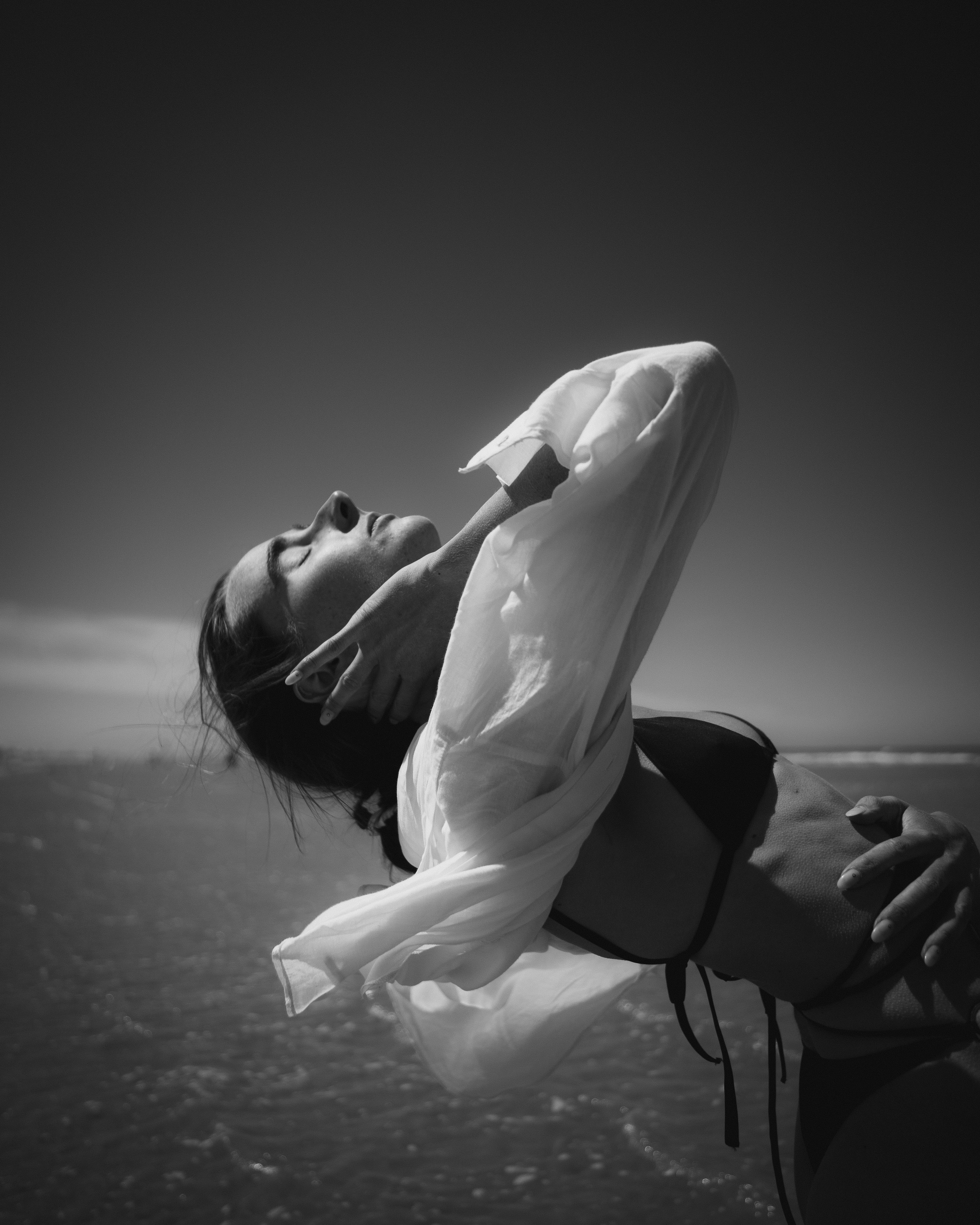 A woman in a bikini and flowing white shirt poses gracefully against a beach backdrop, capturing a moment of serene beauty. The monochrome tones enhance the emotional depth of the scene.