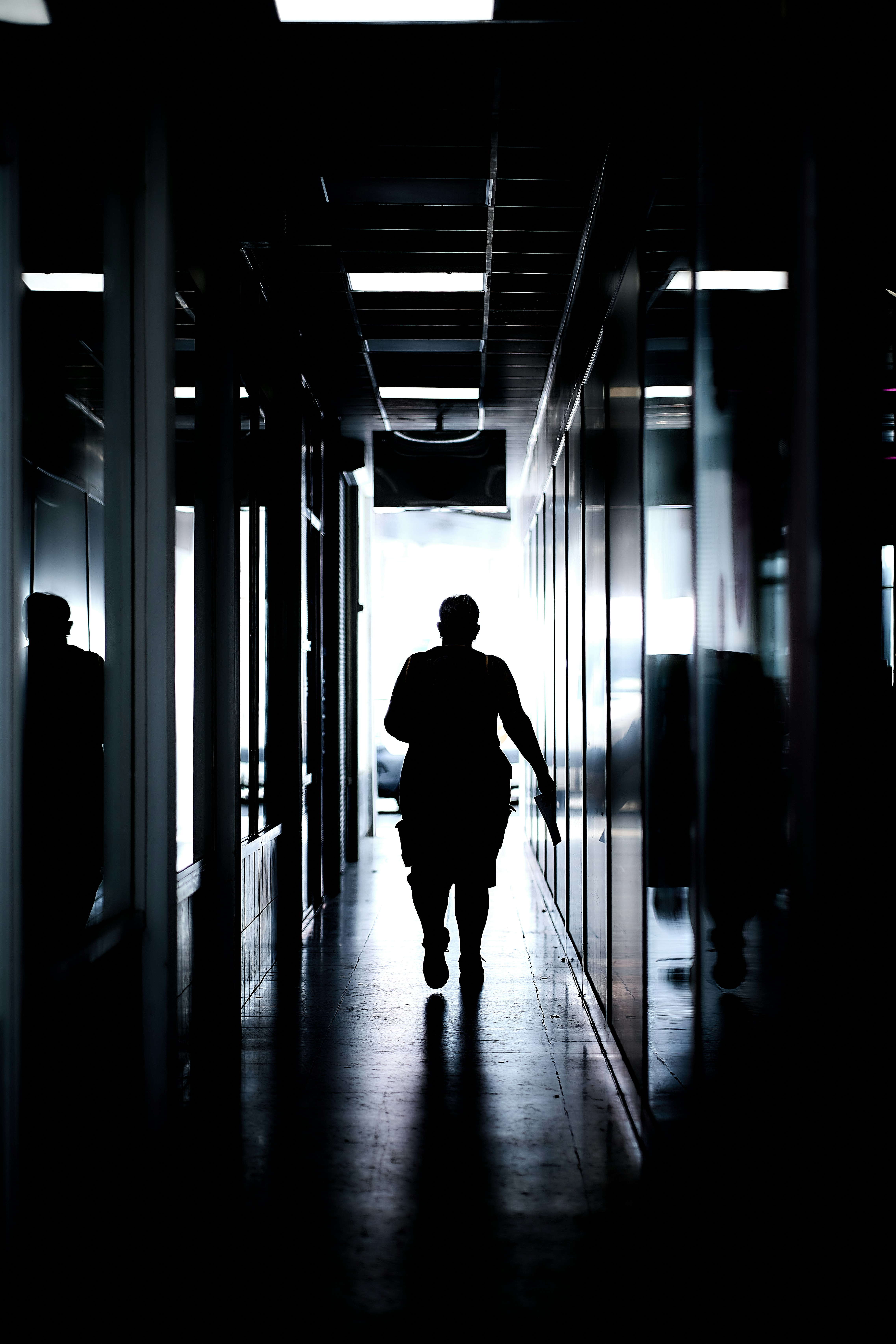 Silhouette of a person walking through a dimly lit corridor, with bright light illuminating the exit in the background.