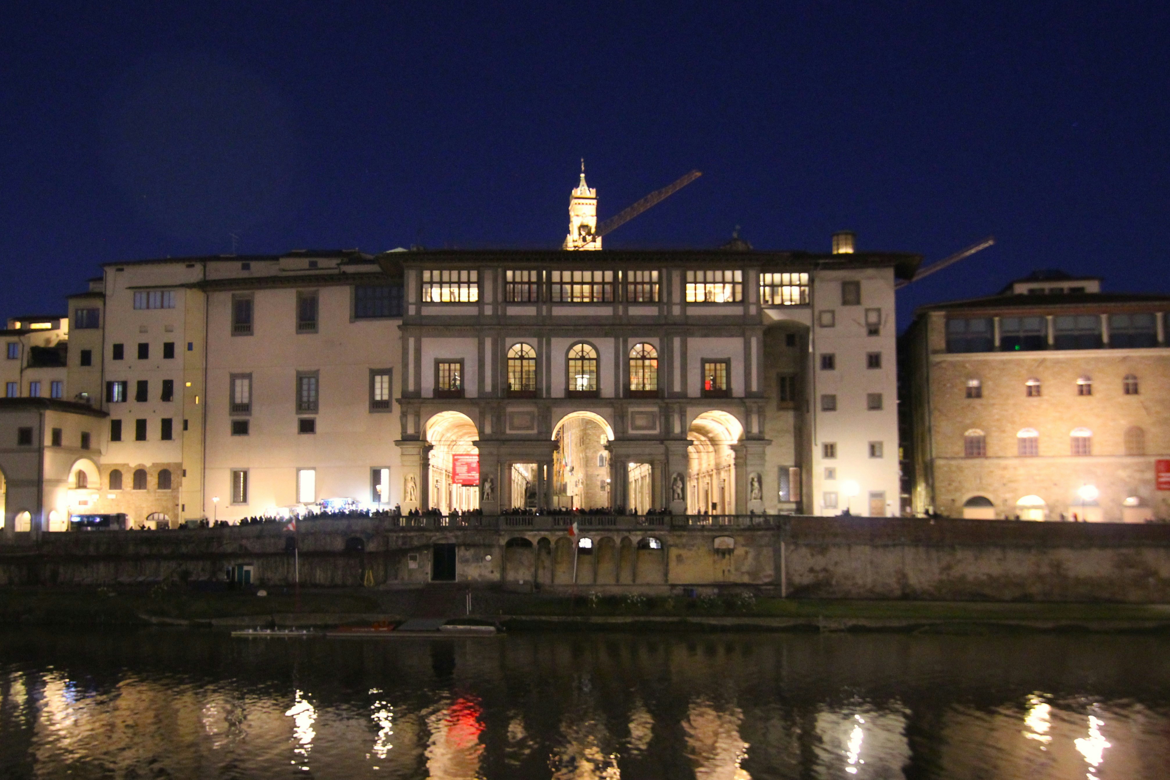 Illuminated Uffizi building with arches reflecting in a calm river under a deep blue twilight sky.
