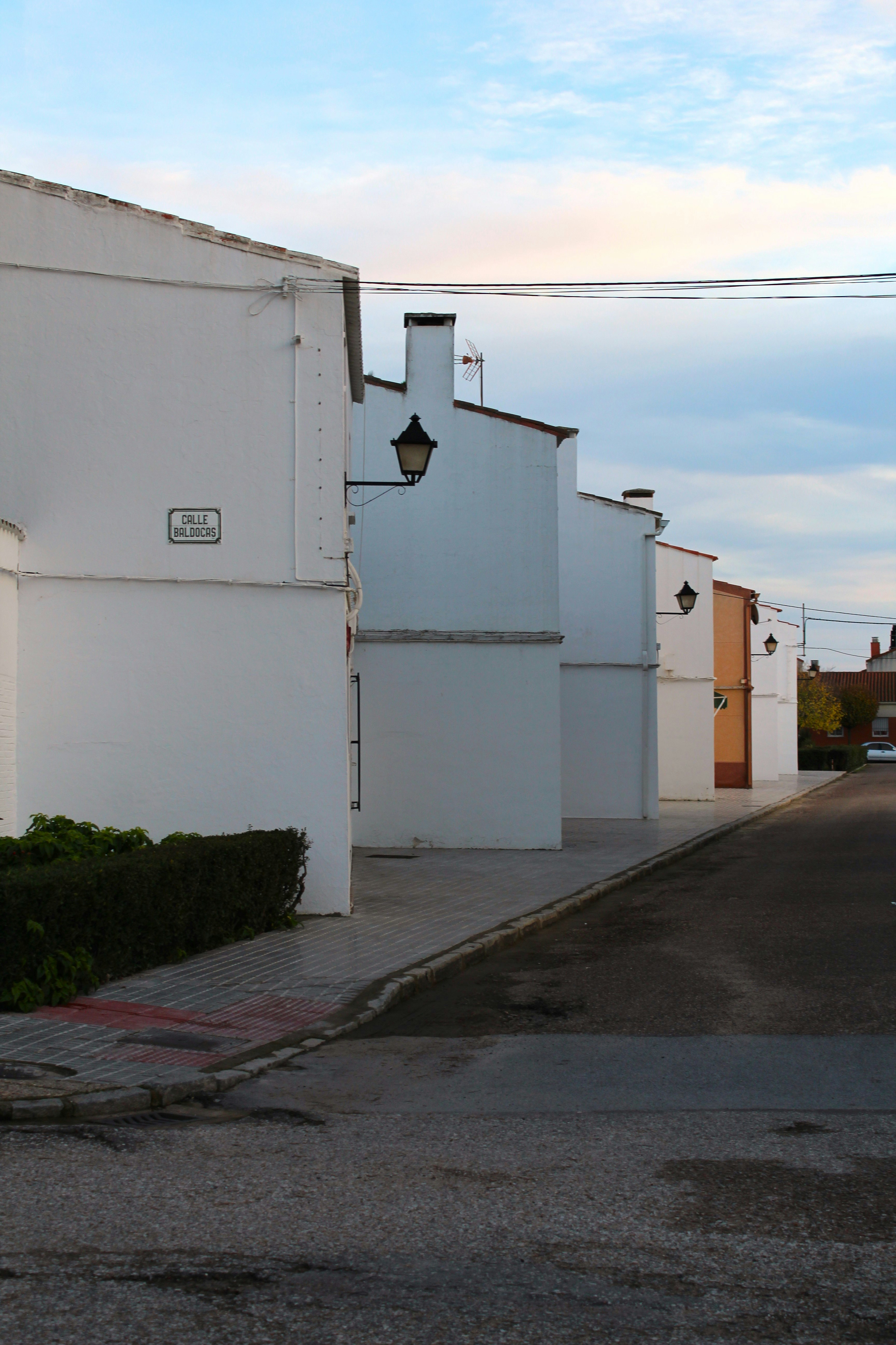 Whitewashed buildings line a tranquil street, illuminated by vintage street lamps under a pastel sky.