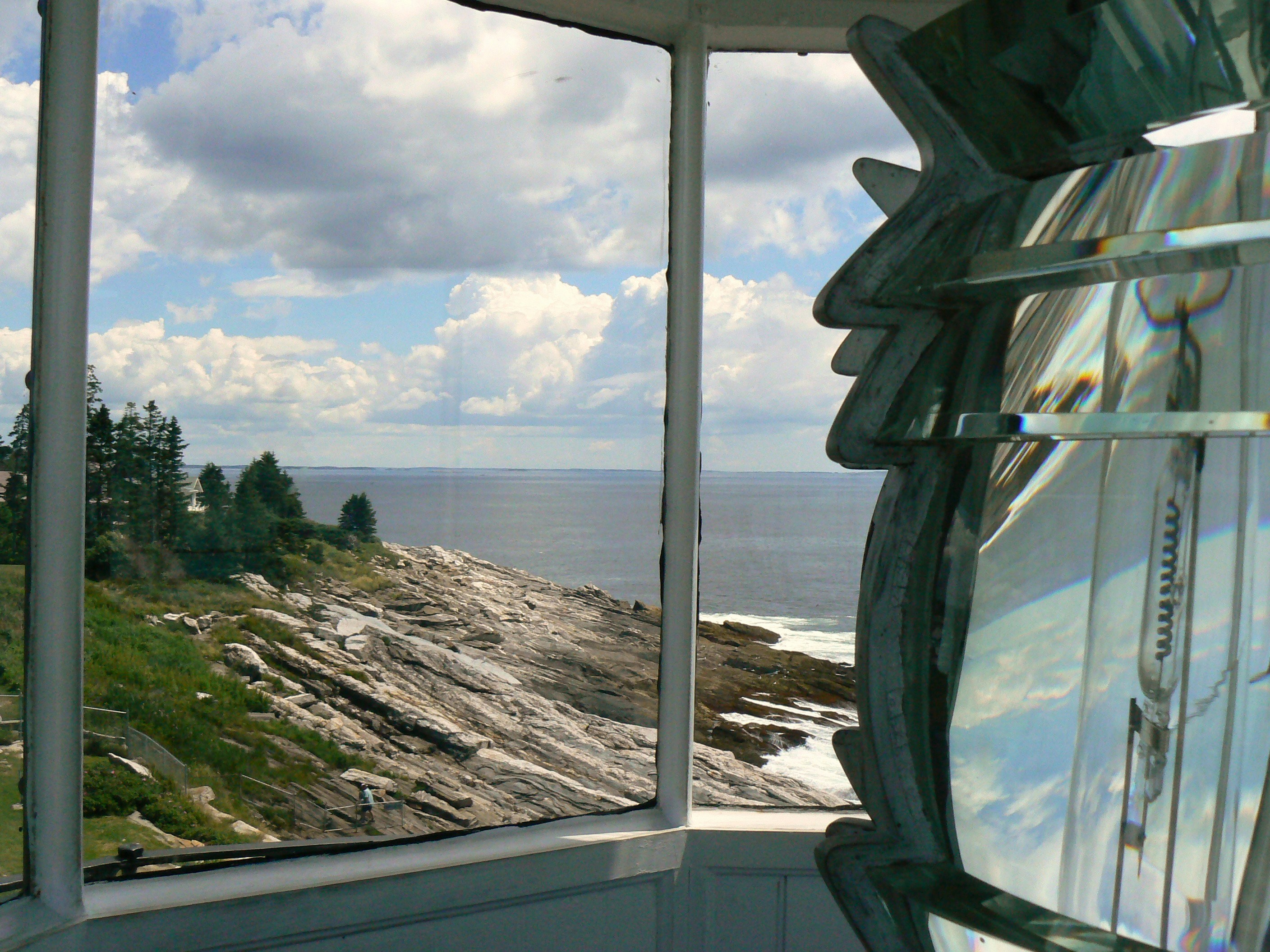 View from a lighthouse showcasing rocky coastline and calm waters under a cloudy sky, with a large lens reflecting the scene.