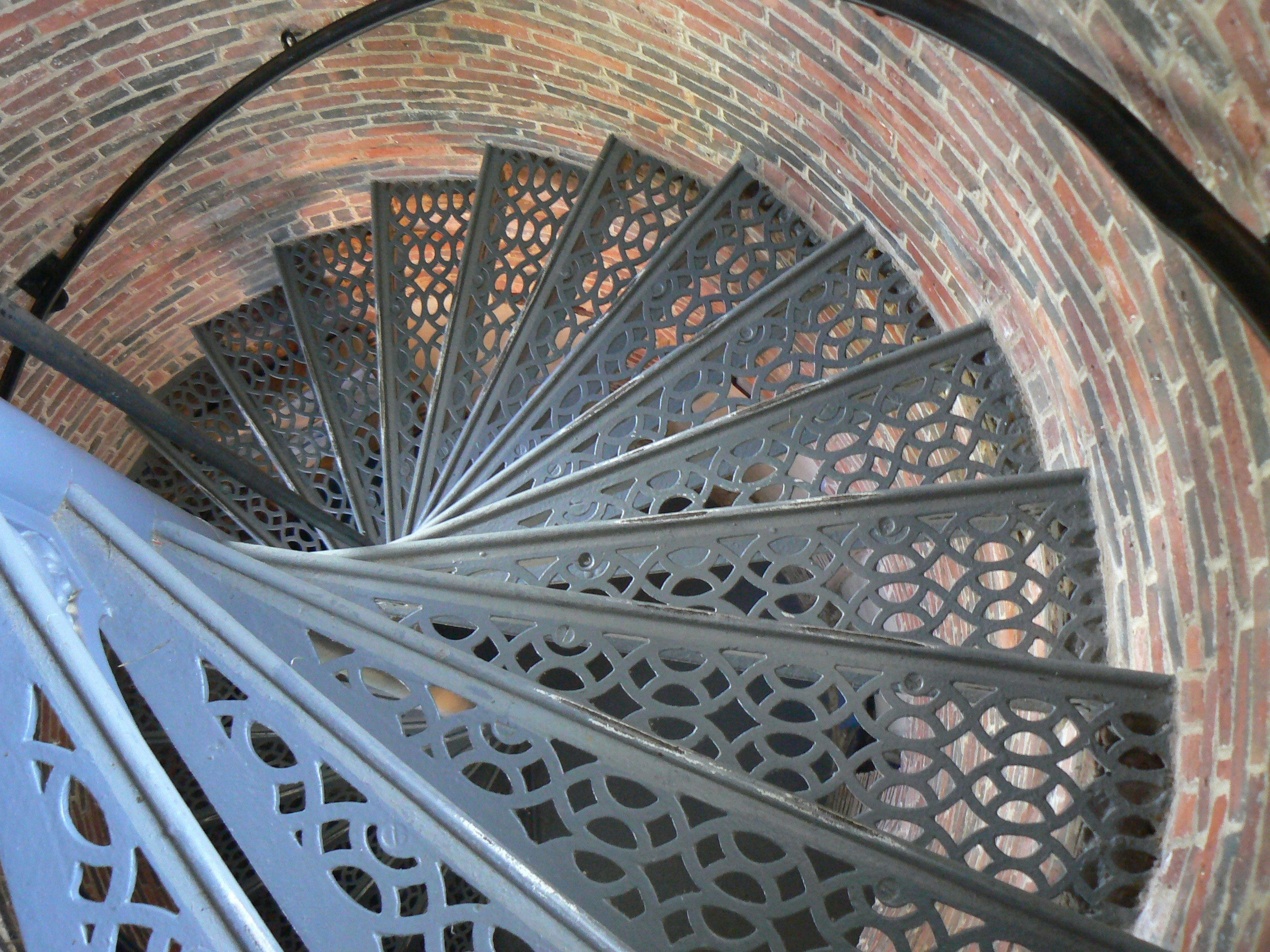 Photograph of a metal spiral staircase winding through a circular brick well, its perforated steps creating a geometric pattern.
