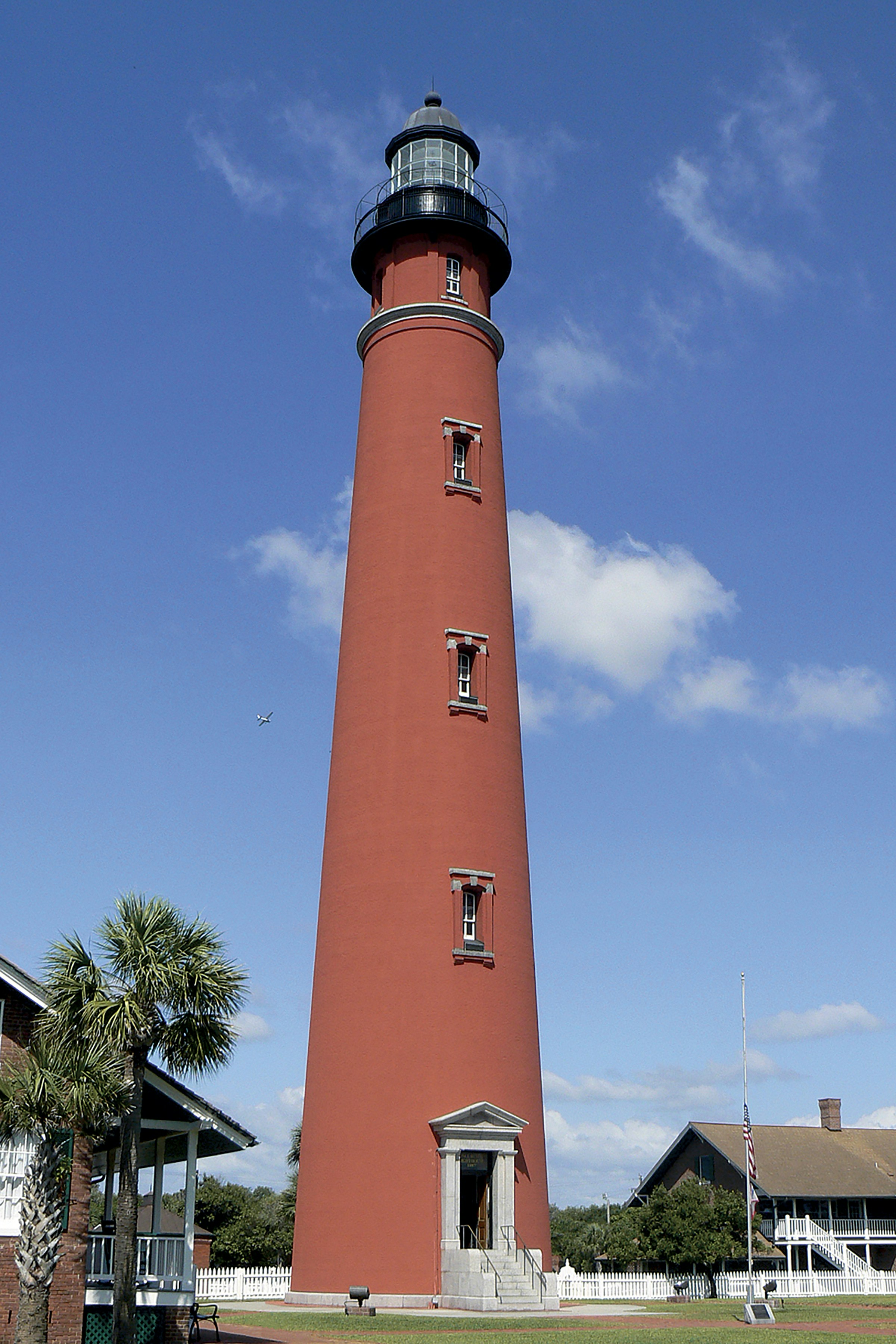 Tall red lighthouse standing against a clear blue sky, surrounded by lush greenery and historic buildings.