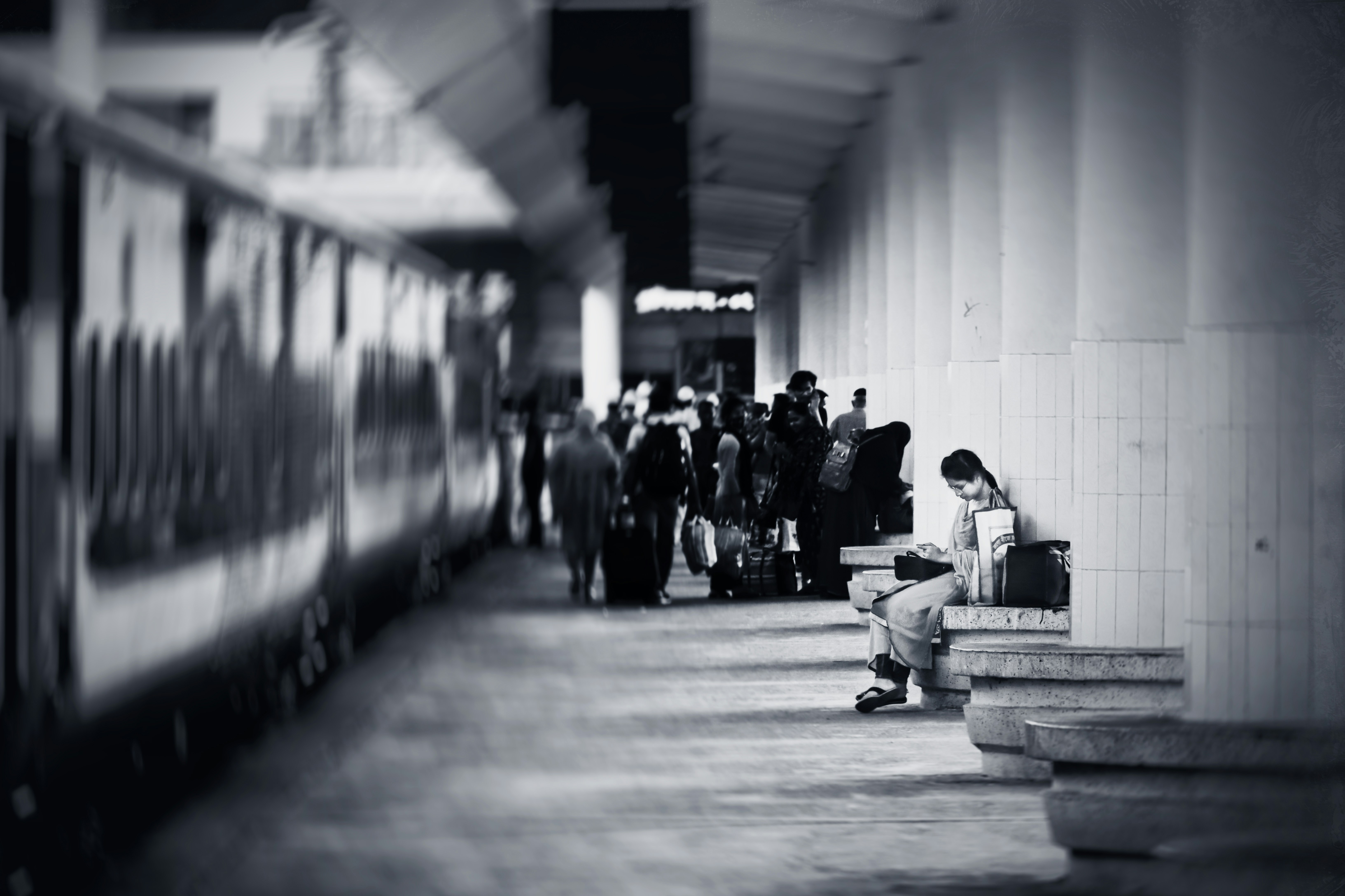 Person seated on a railway platform bench, surrounded by a blurred crowd and train in motion.