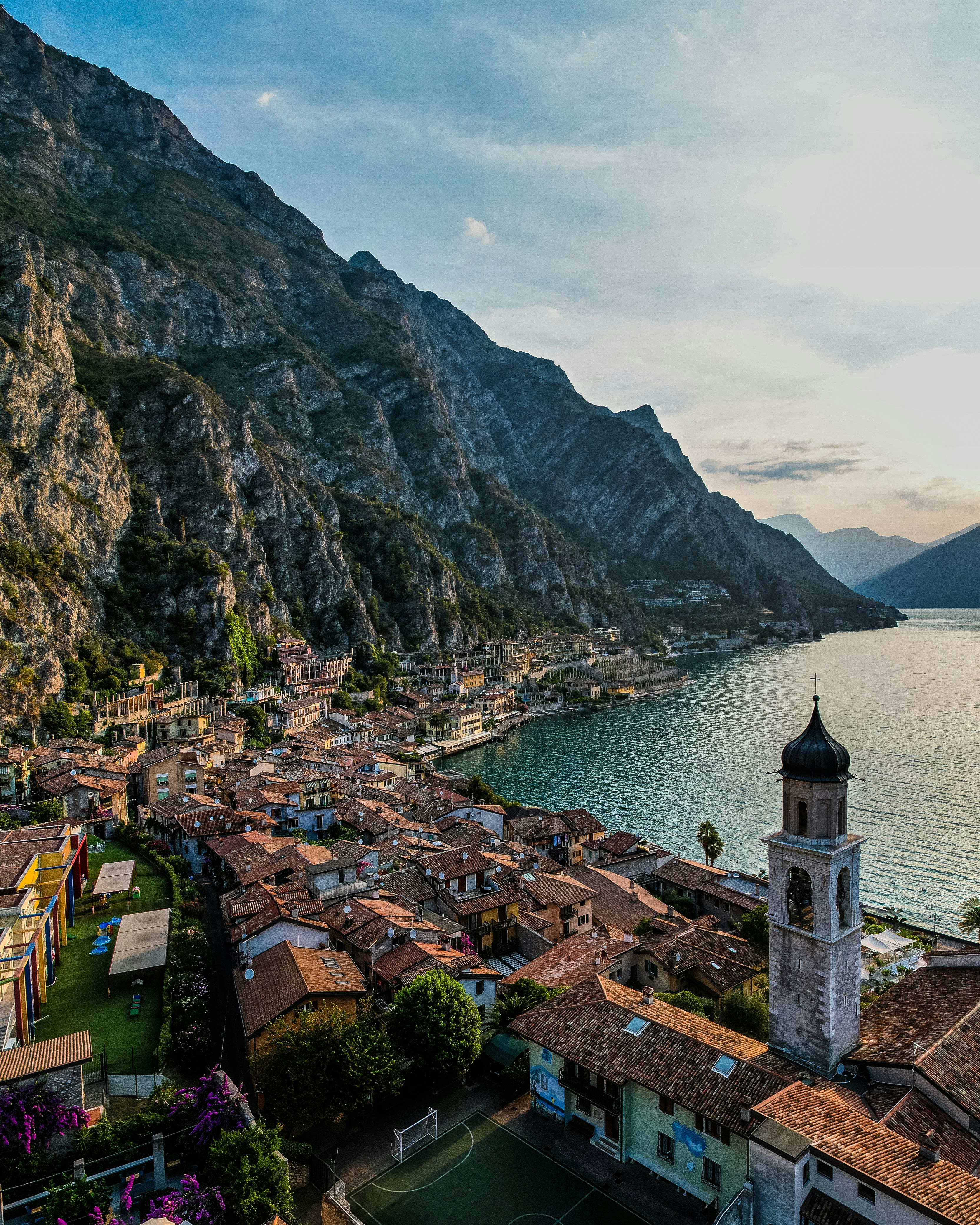 Quaint village nestled along the shores of Lago di Garda, surrounded by majestic mountains and shimmering waters. The scene features charming rooftops and a prominent clock tower.