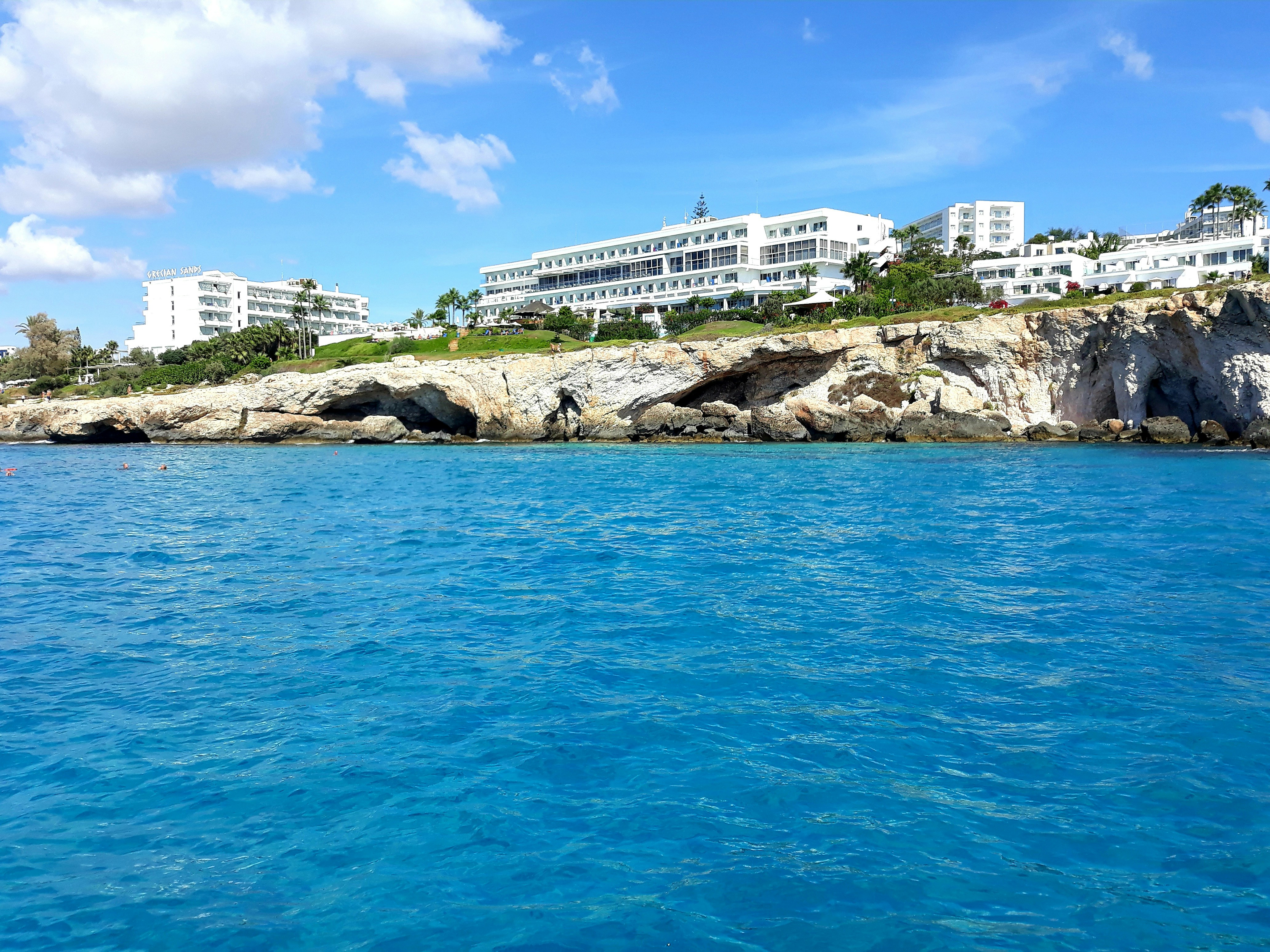 Bright blue ocean with rocky cliffs and white buildings under a partly cloudy sky.
