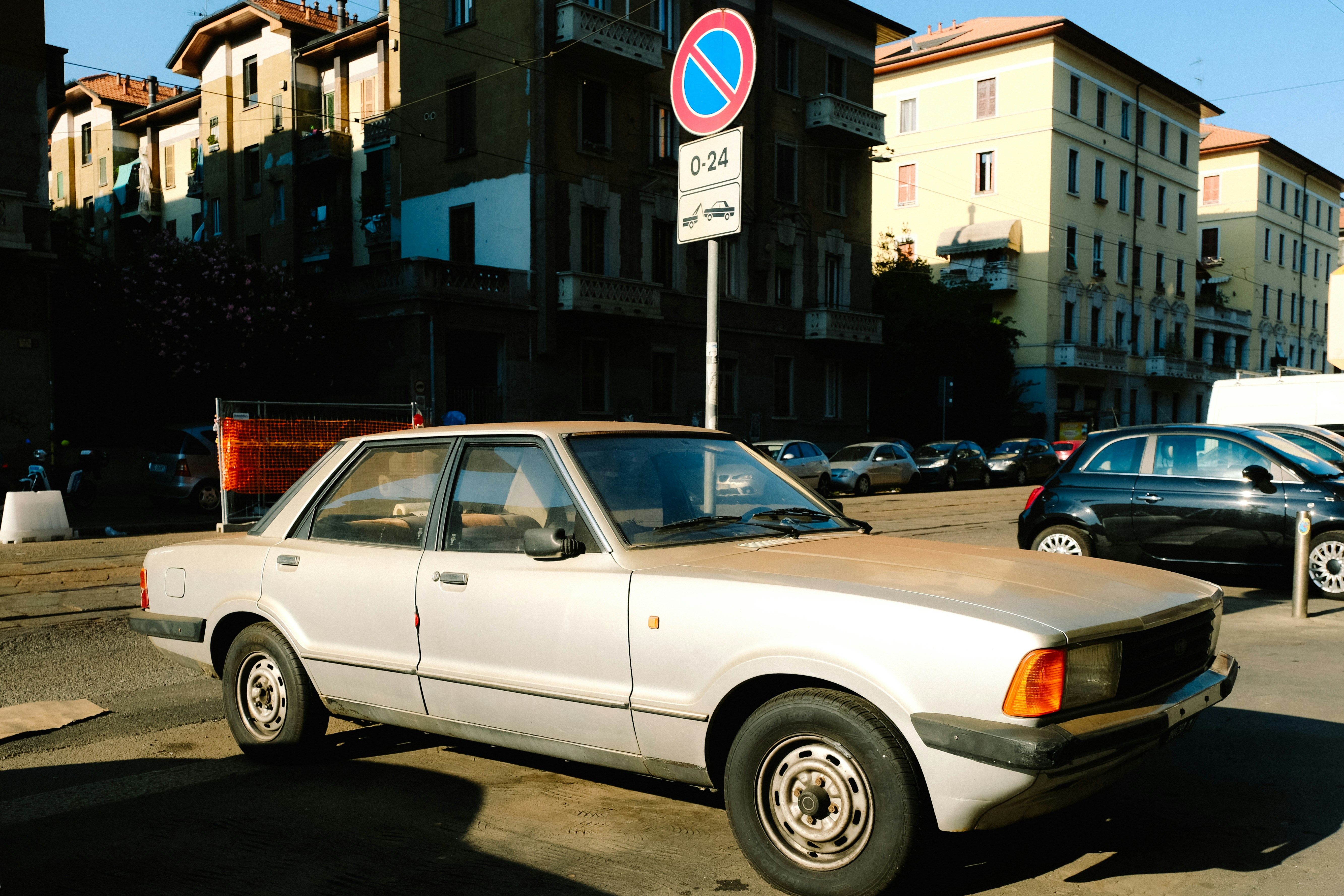 Weathered silver sedan parked on a city street, surrounded by vintage architecture and modern cars. A no-parking sign adds to the urban scene.