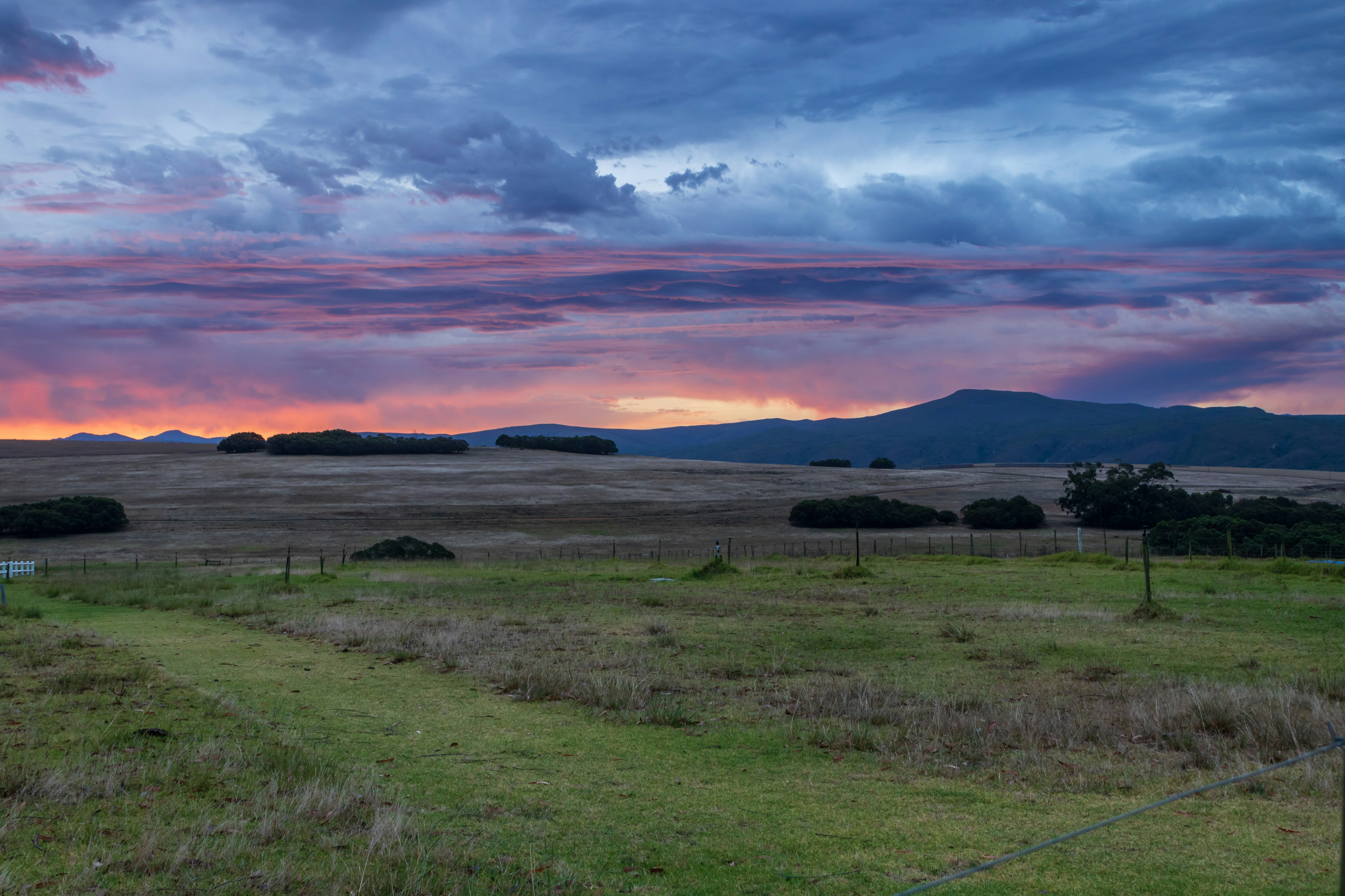 Vibrant twilight sky painted with hues of purple and orange above expansive fields and distant hills. The serene landscape invites reflection.