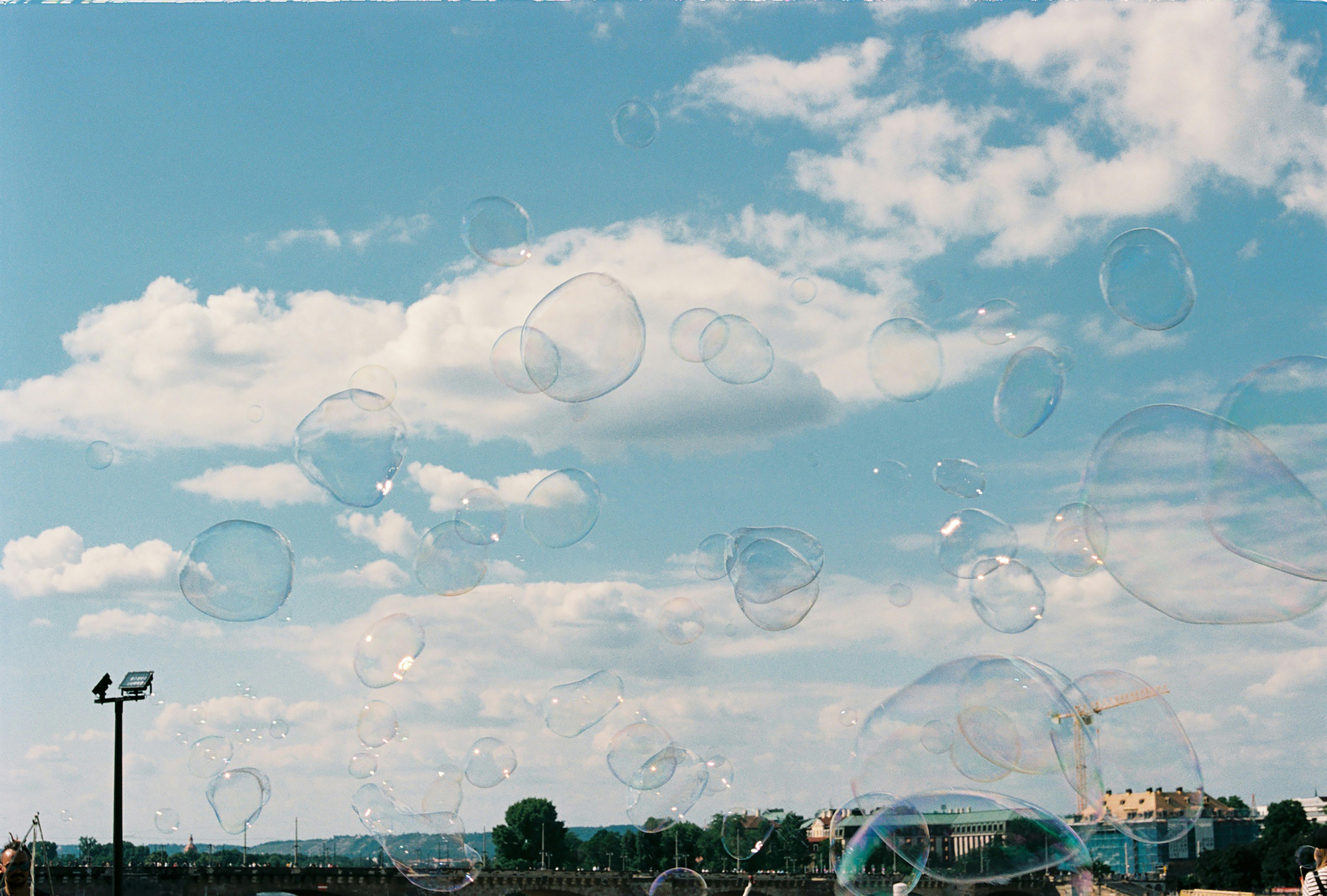 Glistening soap bubbles float against a backdrop of fluffy clouds and a distant cityscape. The scene captures a moment of whimsy and joy.
