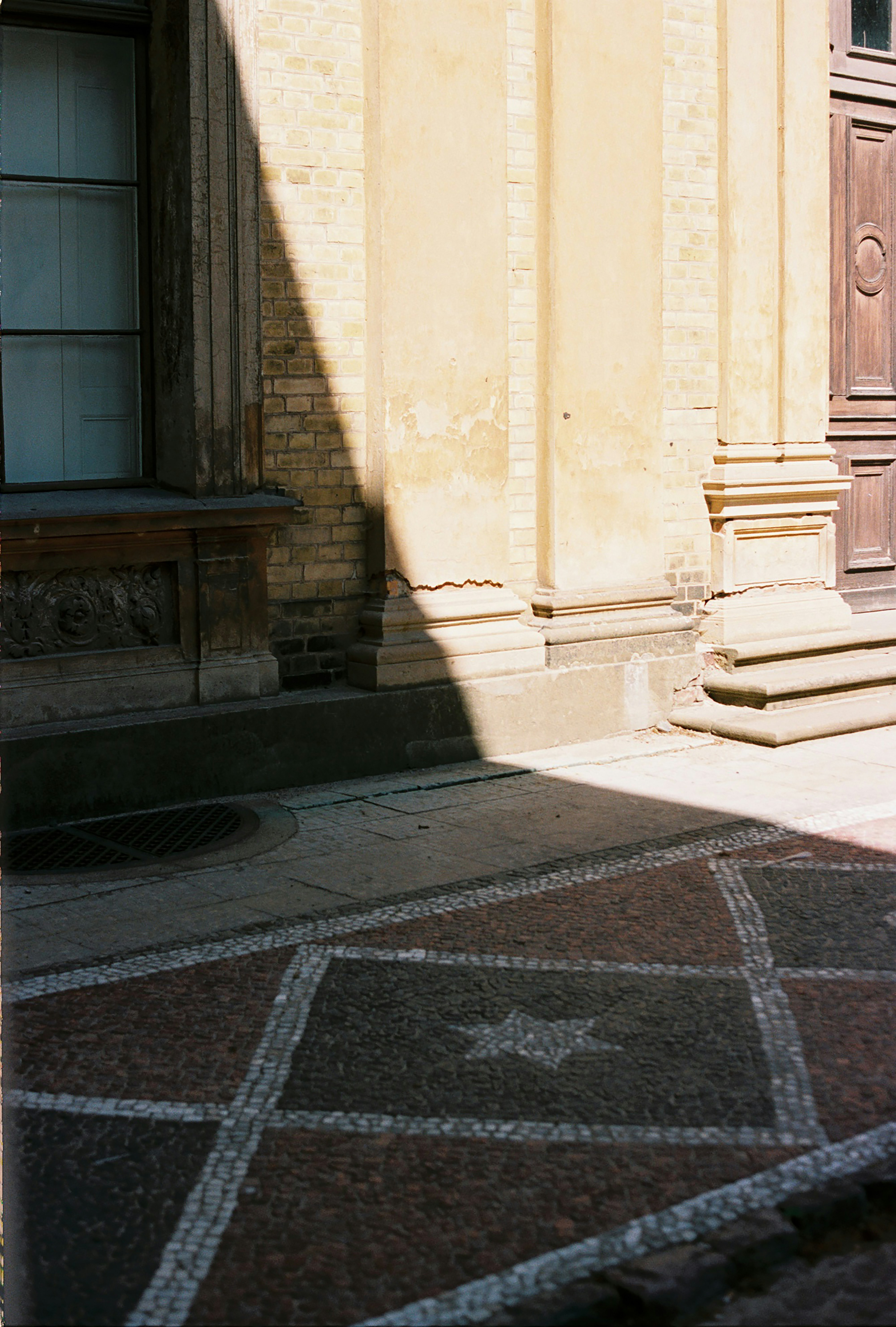 Sunlit facade of a historic building with tall columns and carved stonework. The star-patterned pavement sits in diagonal shadow, emphasizing geometric detail.