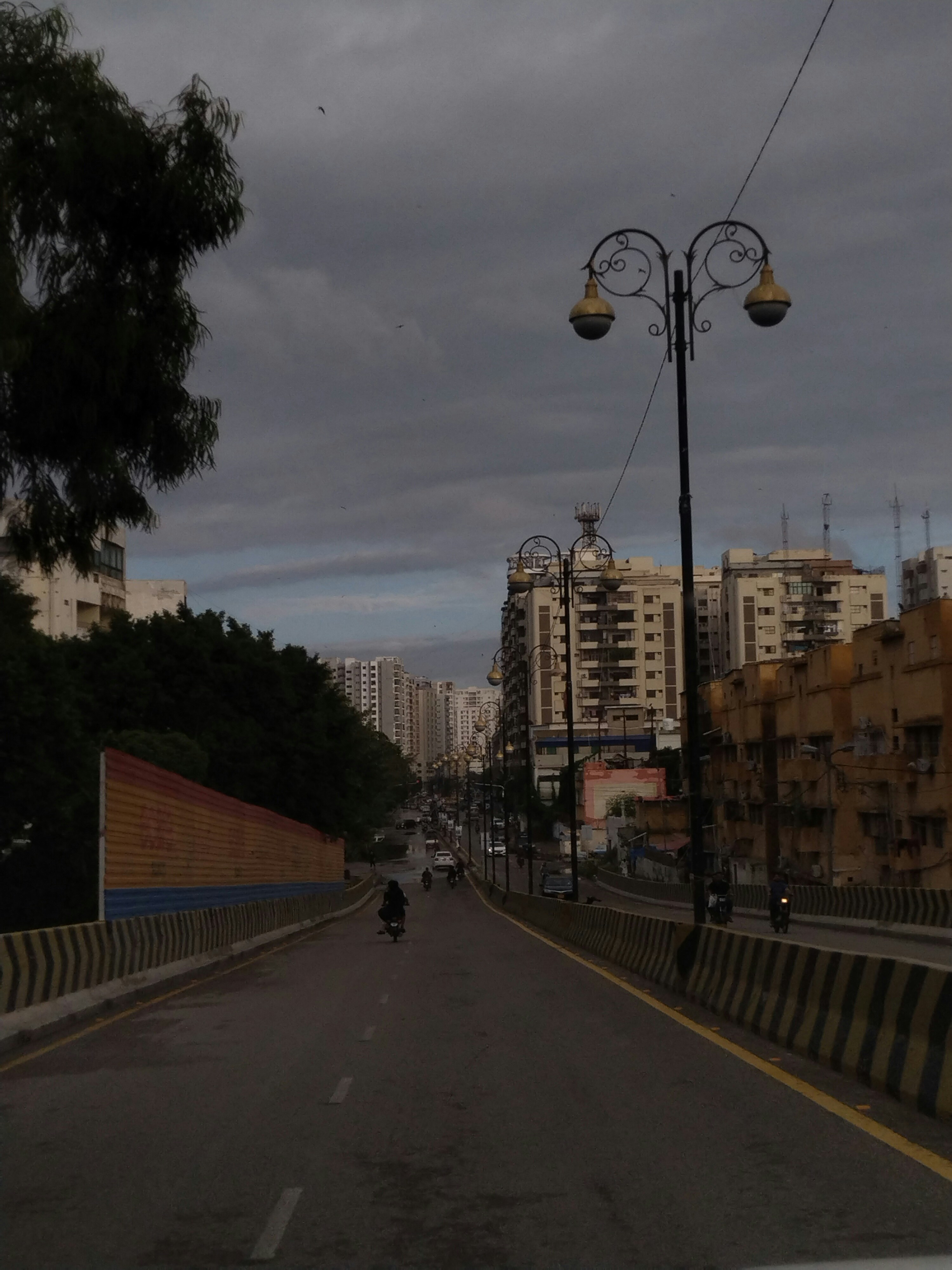 A city road flanked by tall buildings and street lamps under a moody sky. Motorcycles navigate the quiet street, hinting at urban life.