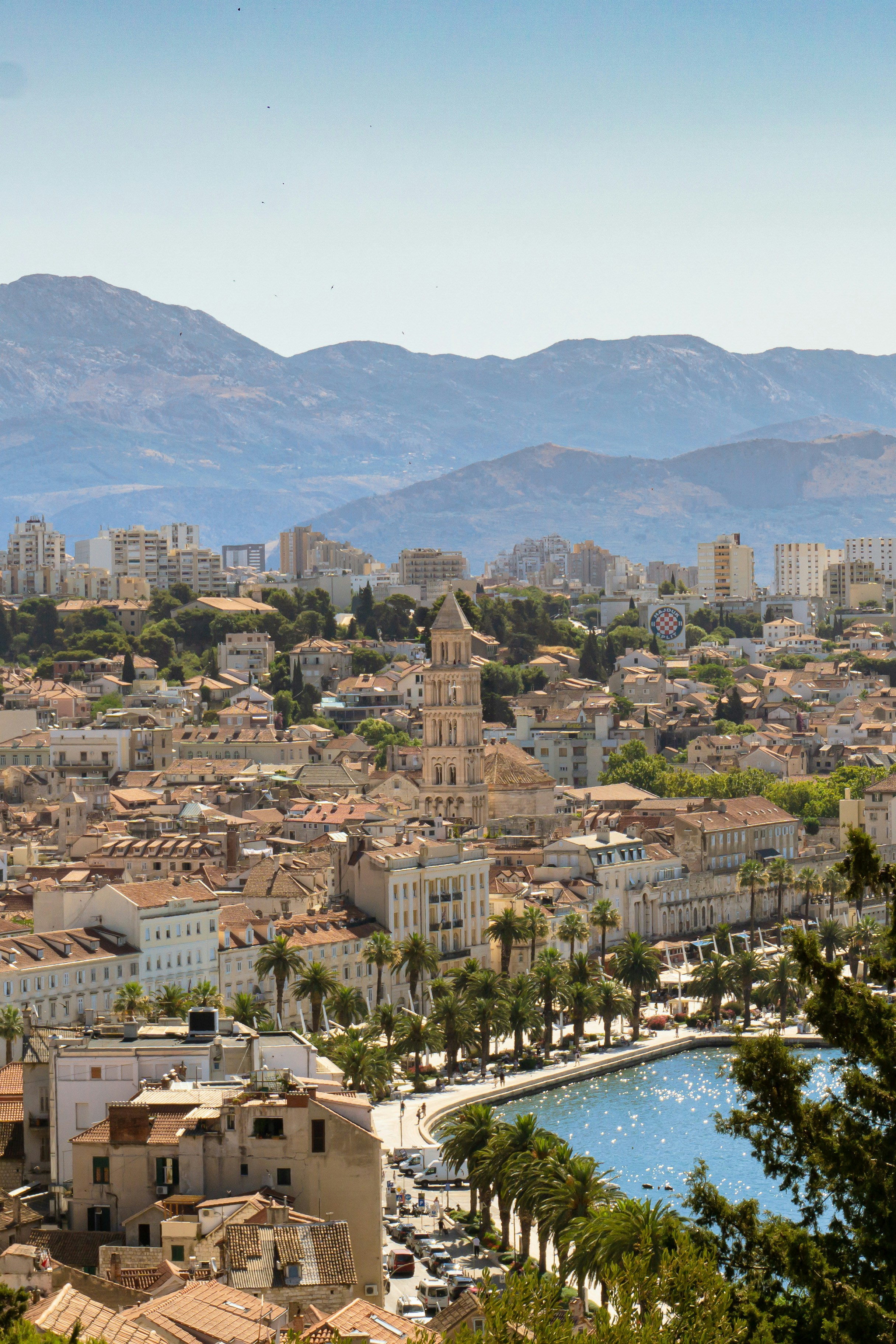 A panoramic view of a coastal city with historic architecture and palm-lined promenades, framed by majestic mountains in the background.