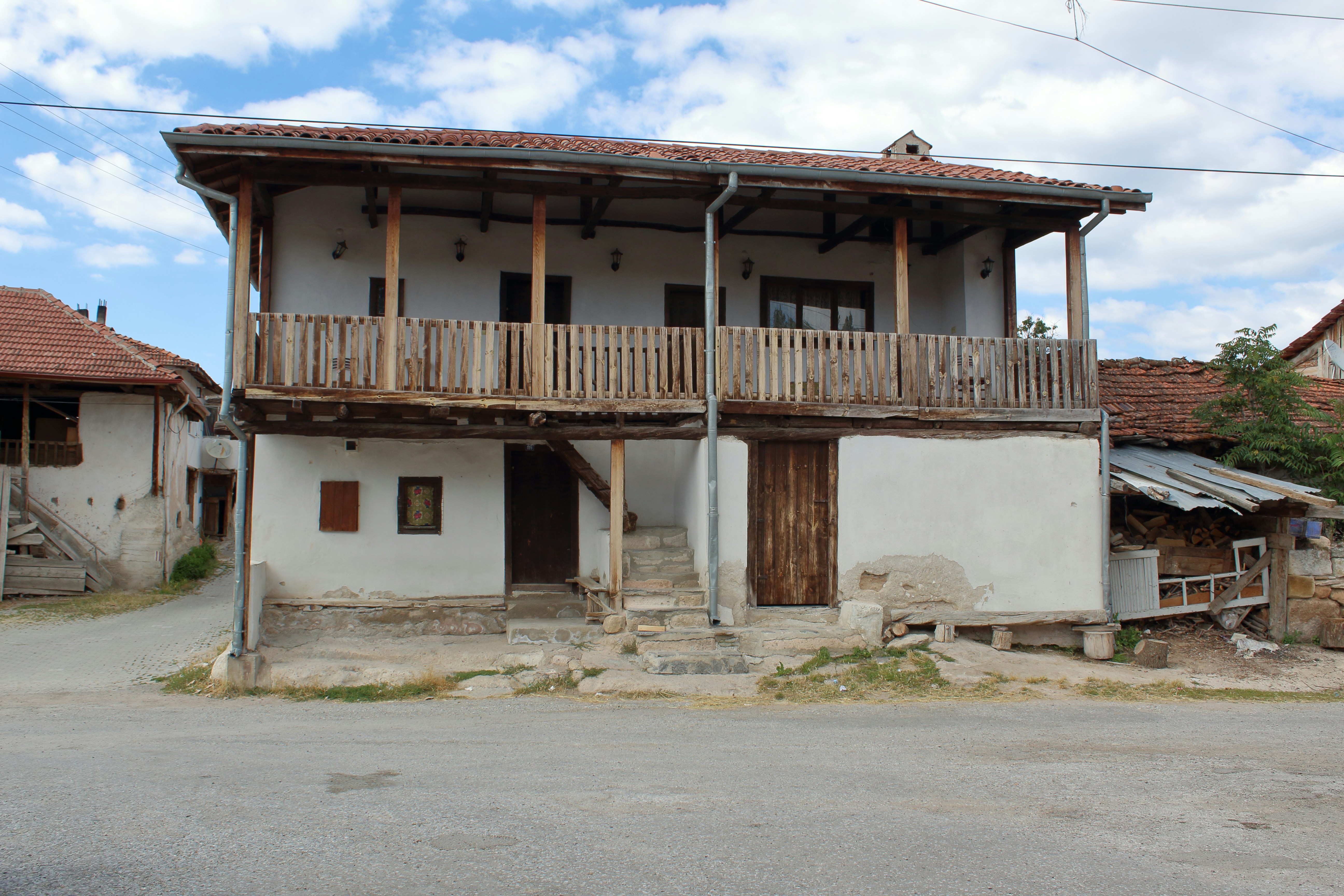 Two-story wooden house with a balcony, set in a rural landscape, showcasing traditional architectural elements. The structure stands against a blue sky dotted with clouds.