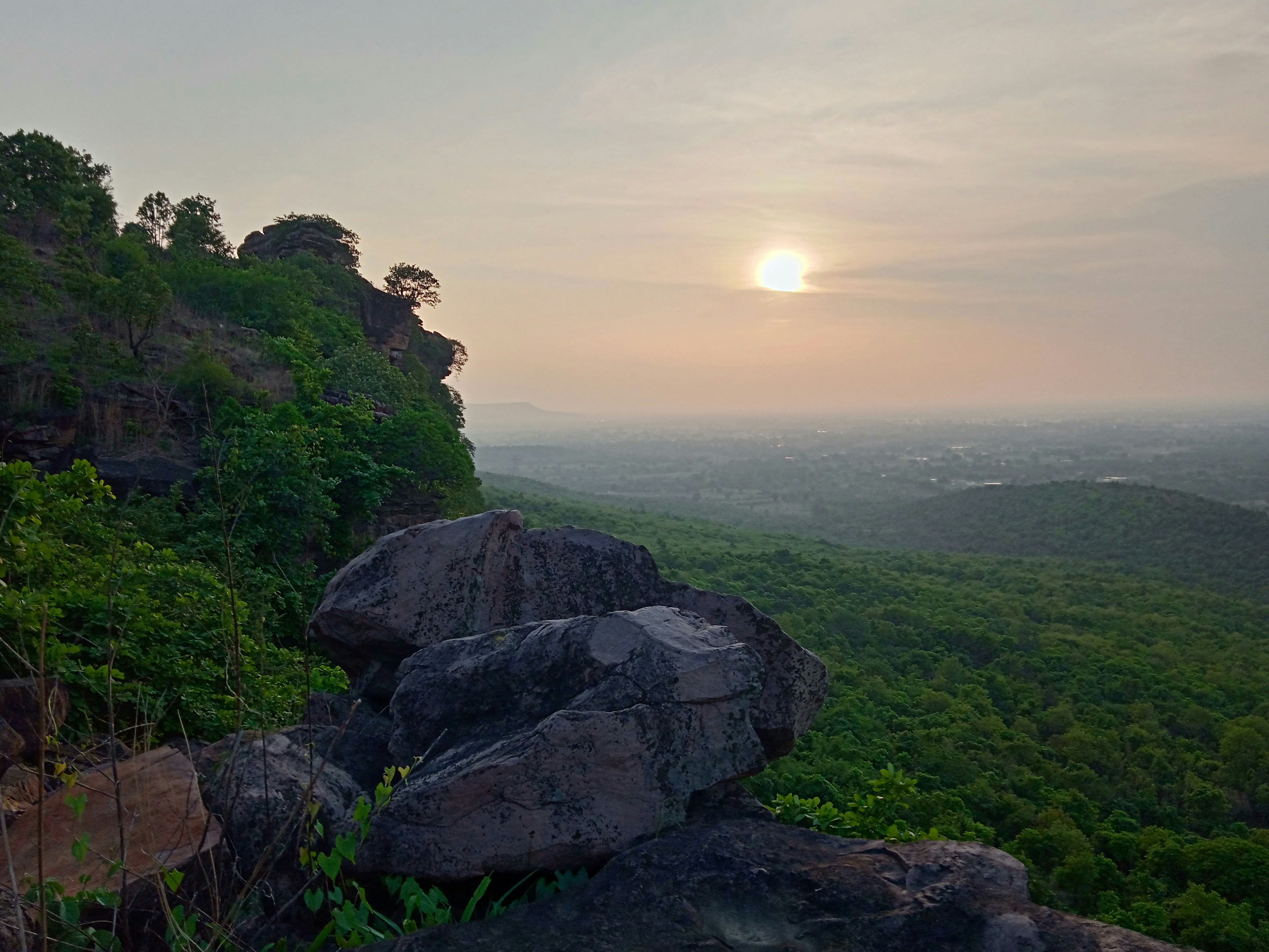 Sunset casting a warm glow over a lush valley framed by rocky outcrops and greenery. The horizon stretches into the distance.
