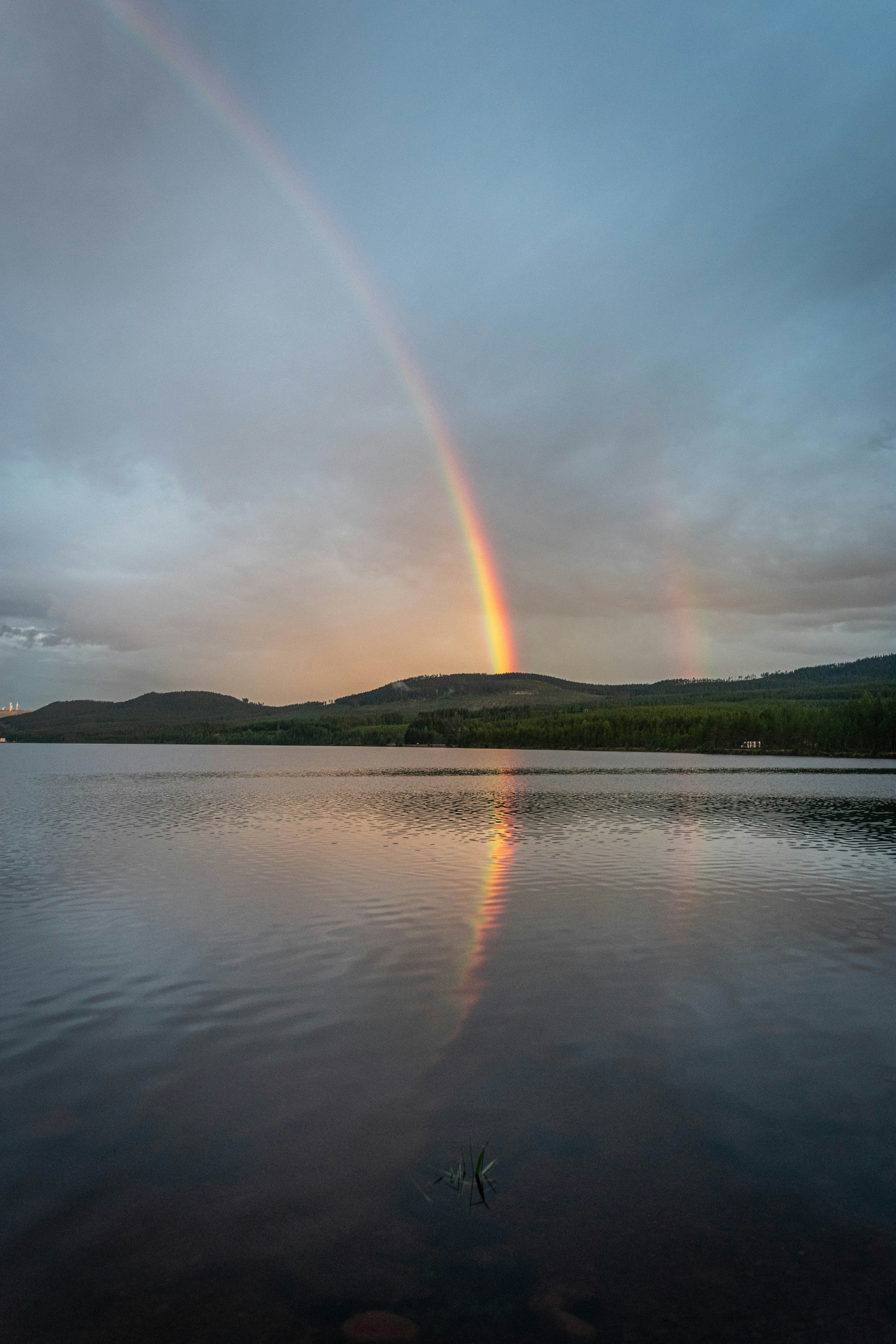 Vibrant rainbow arches over a serene lake against a backdrop of hills and a softly lit sky.