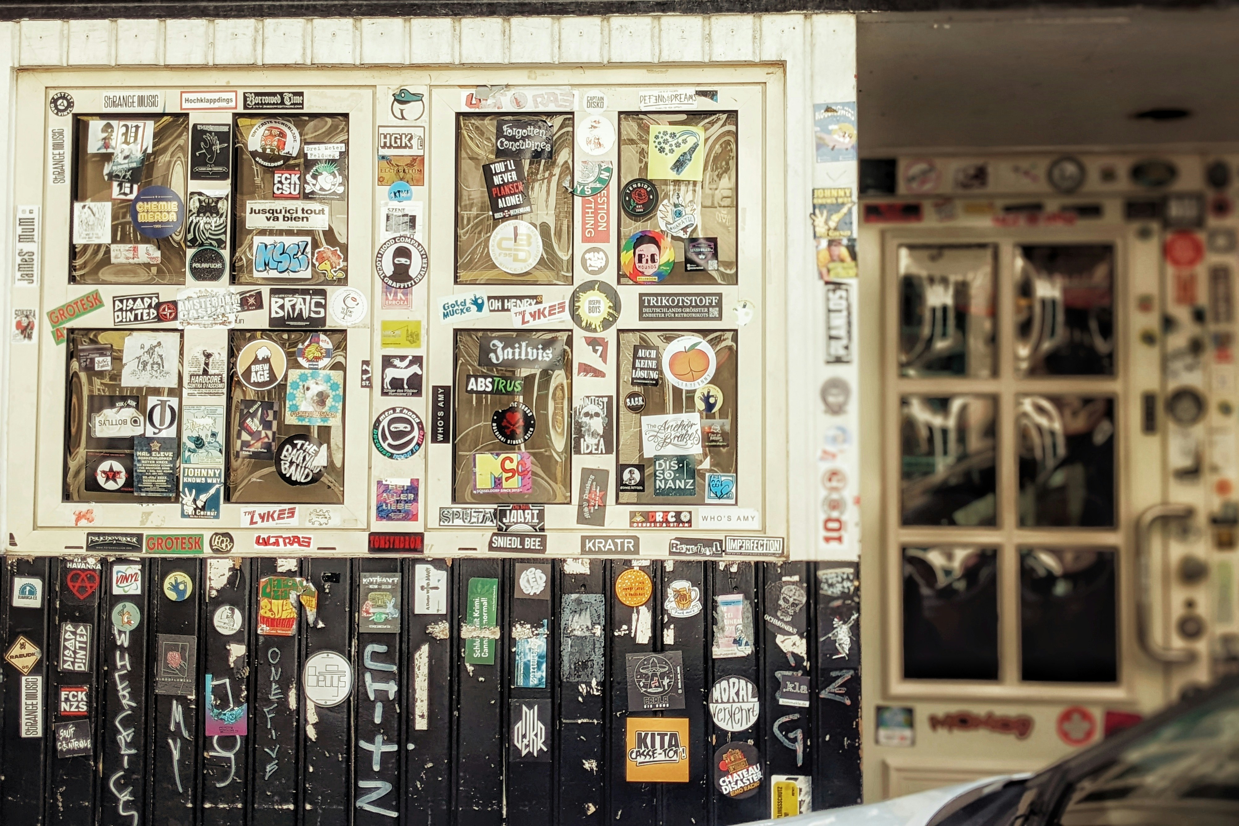 Sticker-covered storefront facade densely packed with posters, logos, and graffiti, with a glass door to the right.