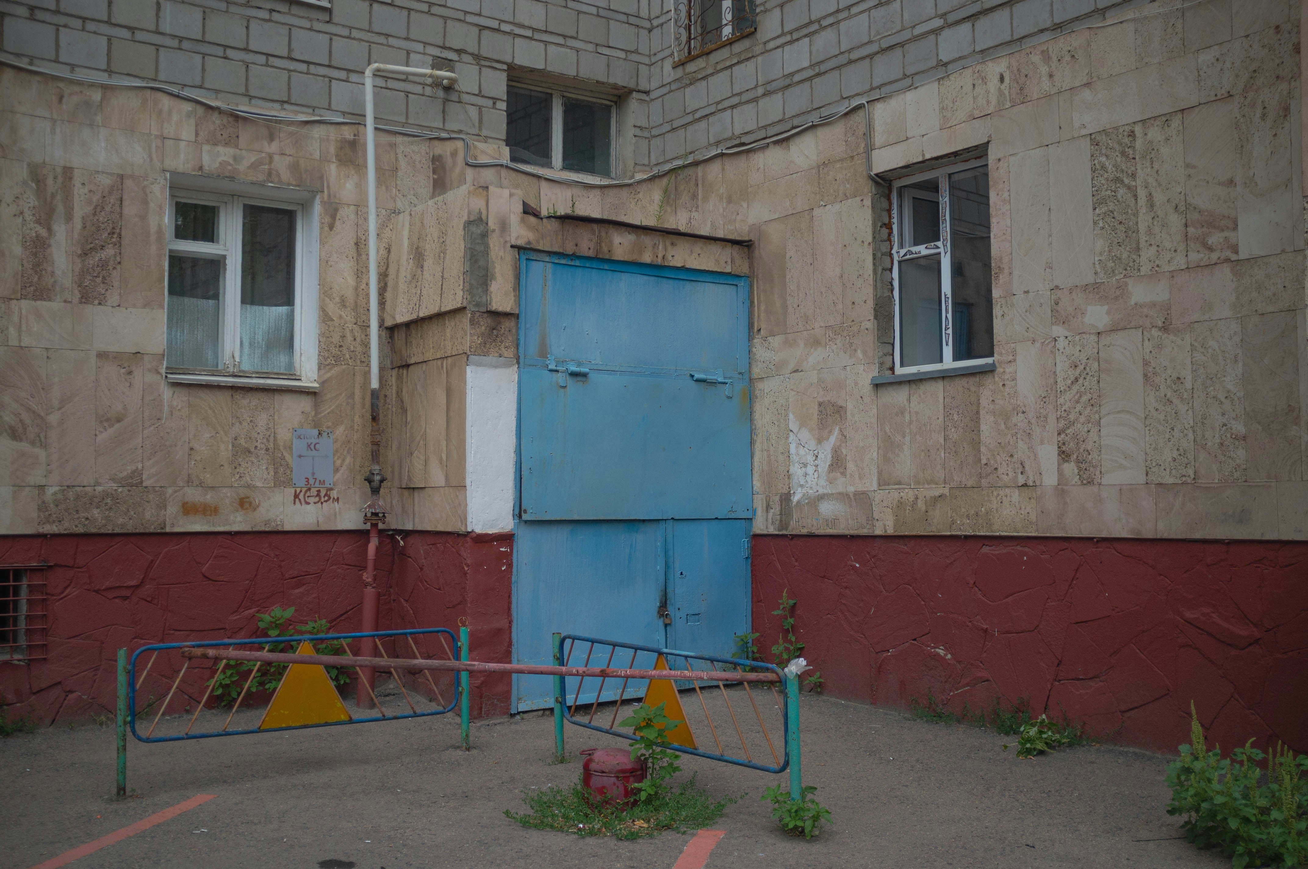 Weathered blue door set against a textured stone wall, surrounded by urban greenery and a barricade. The scene evokes a sense of mystery and history.