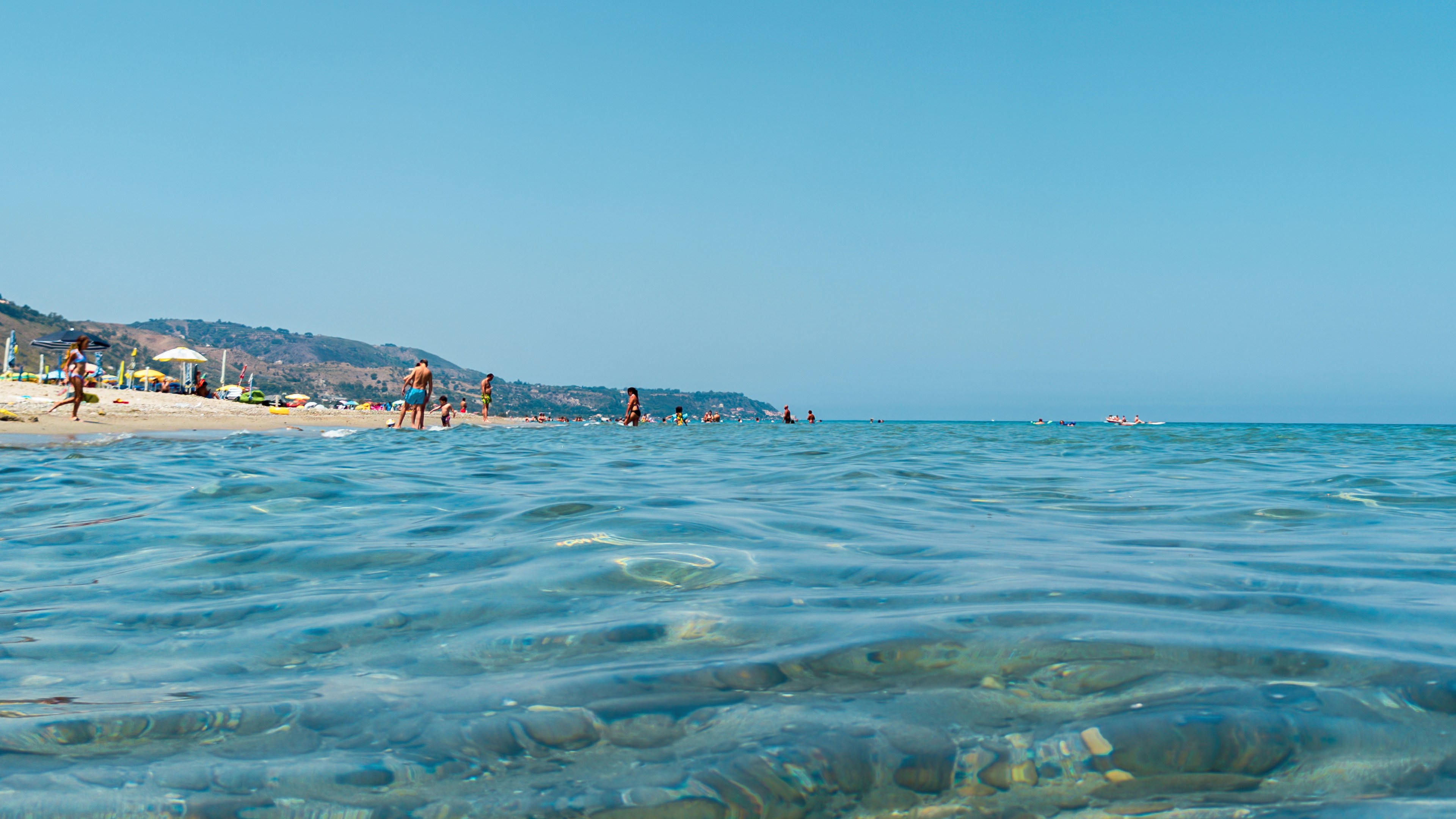 Clear turquoise waters gently lap against a sandy beach, dotted with sunbathers and distant hills under a clear blue sky.