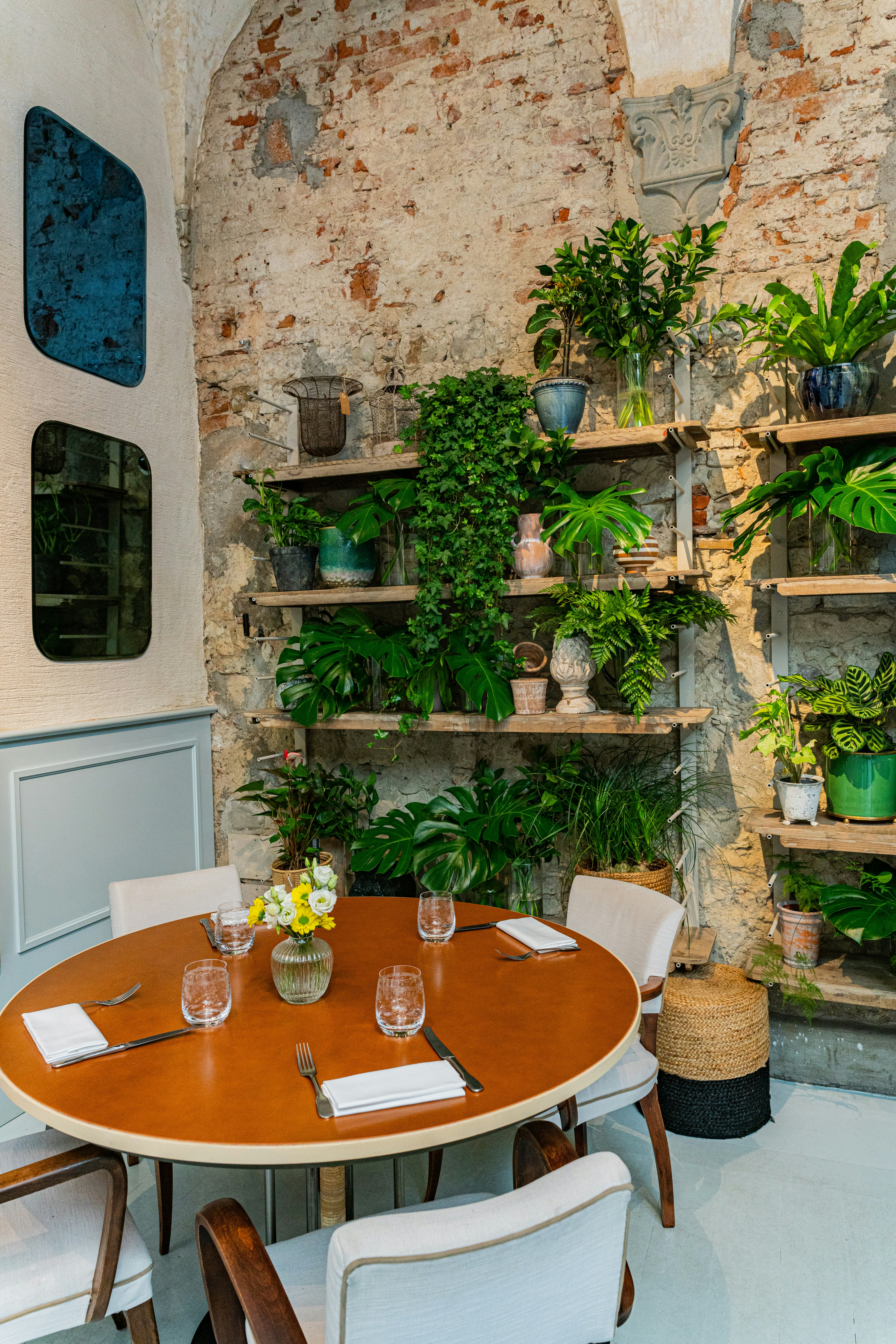 Charming dining area featuring a round table set for a meal, surrounded by lush greenery on wooden shelves against a rustic wall.