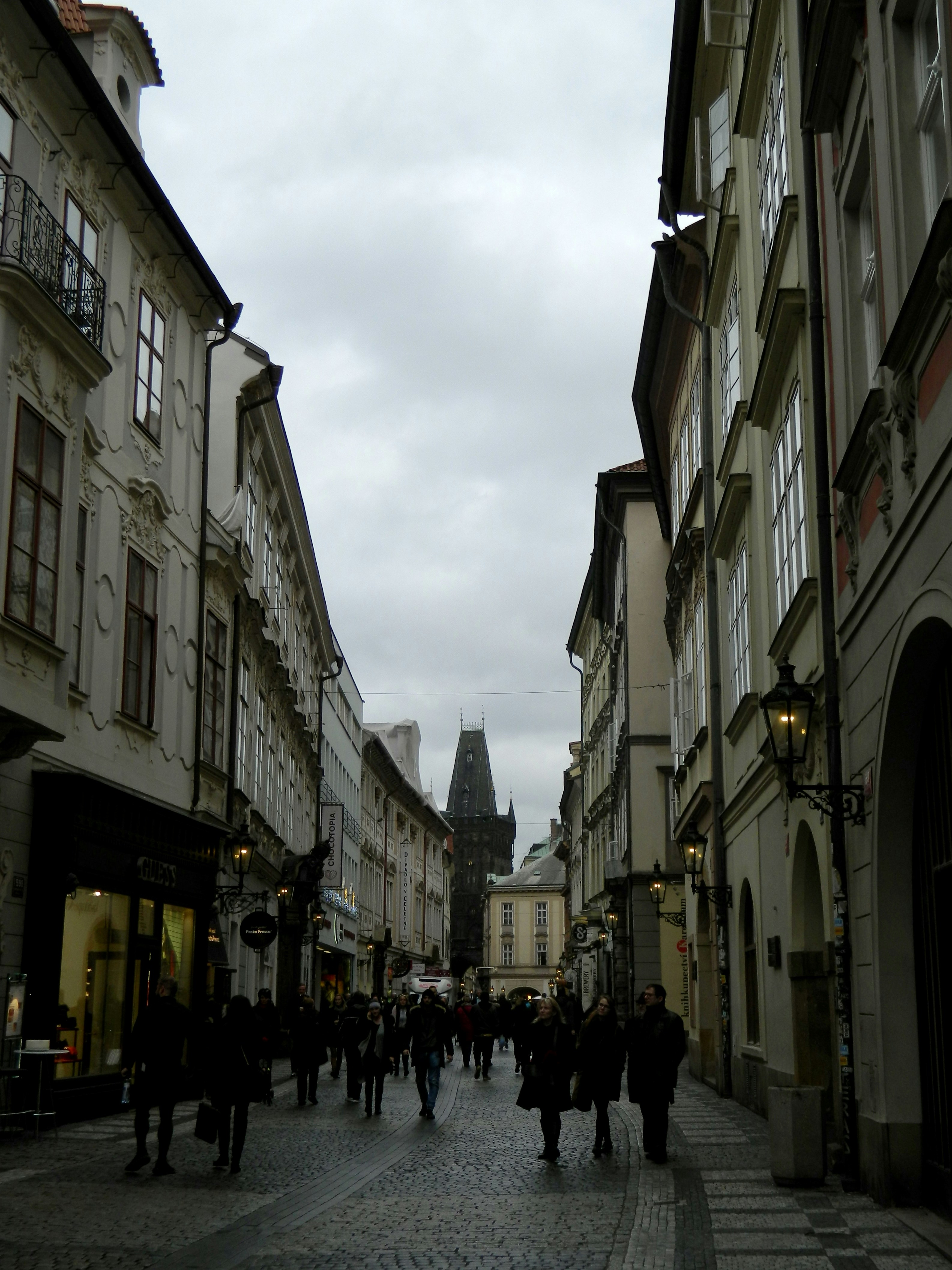 Charming cobblestone street lined with historic buildings, leading to a distant tower under a cloudy sky. Lanterns illuminate the bustling crowd.