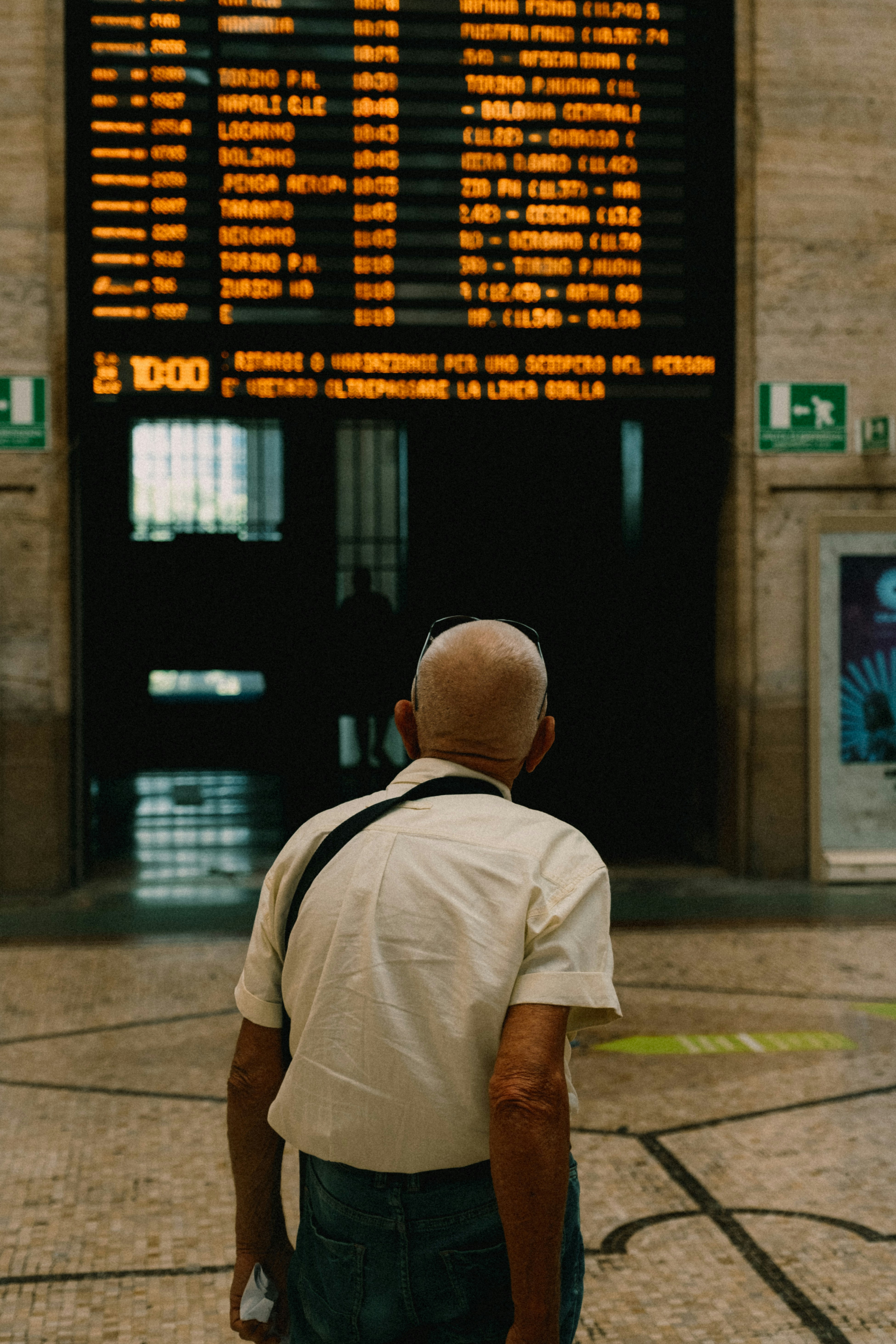 Elderly man gazing at a departure board in a bustling train station, contemplating his next journey. The digital display lists various train schedules.