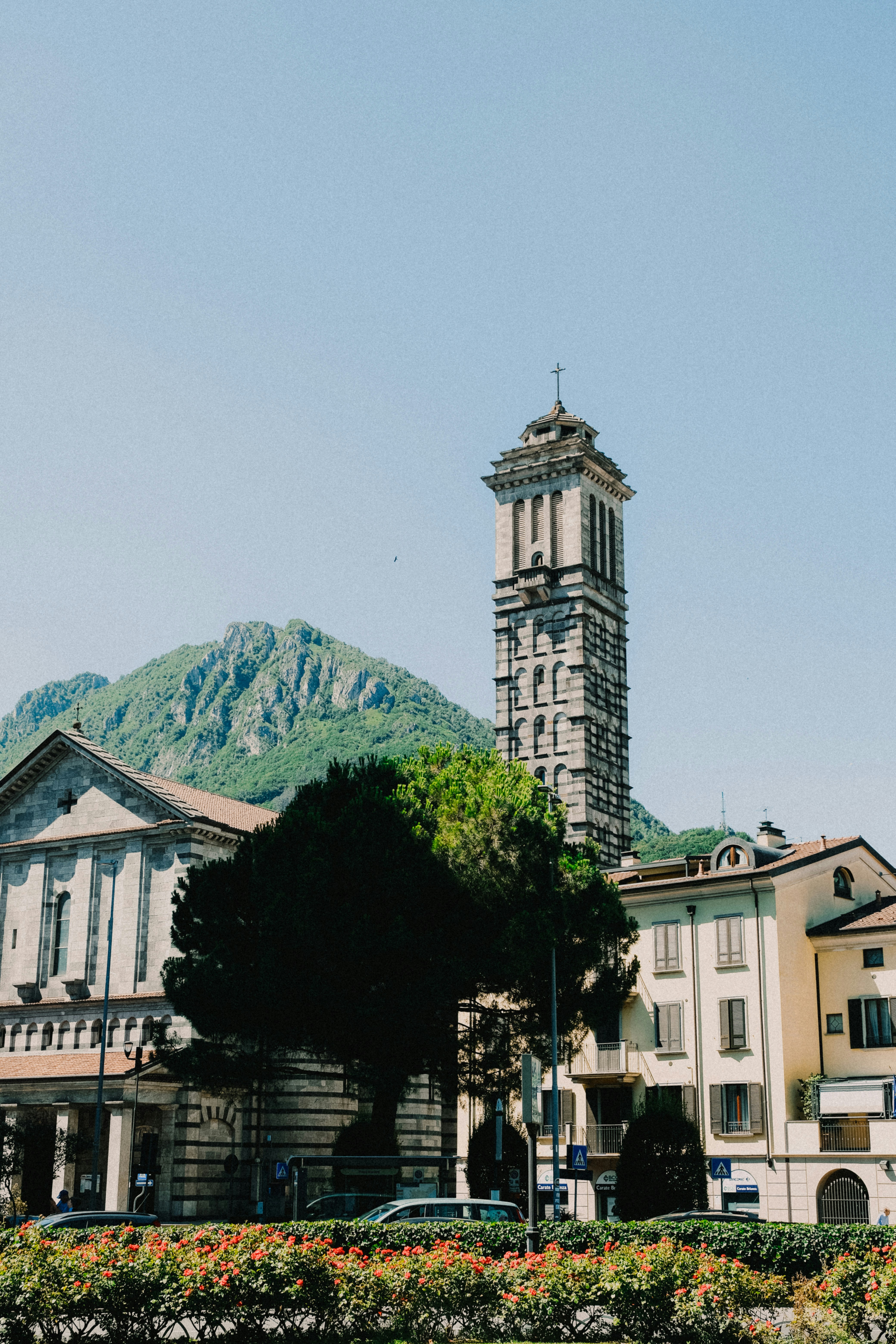 Historic stone bell tower rises above a quaint town, framed by lush mountains and vibrant flowers in the foreground.